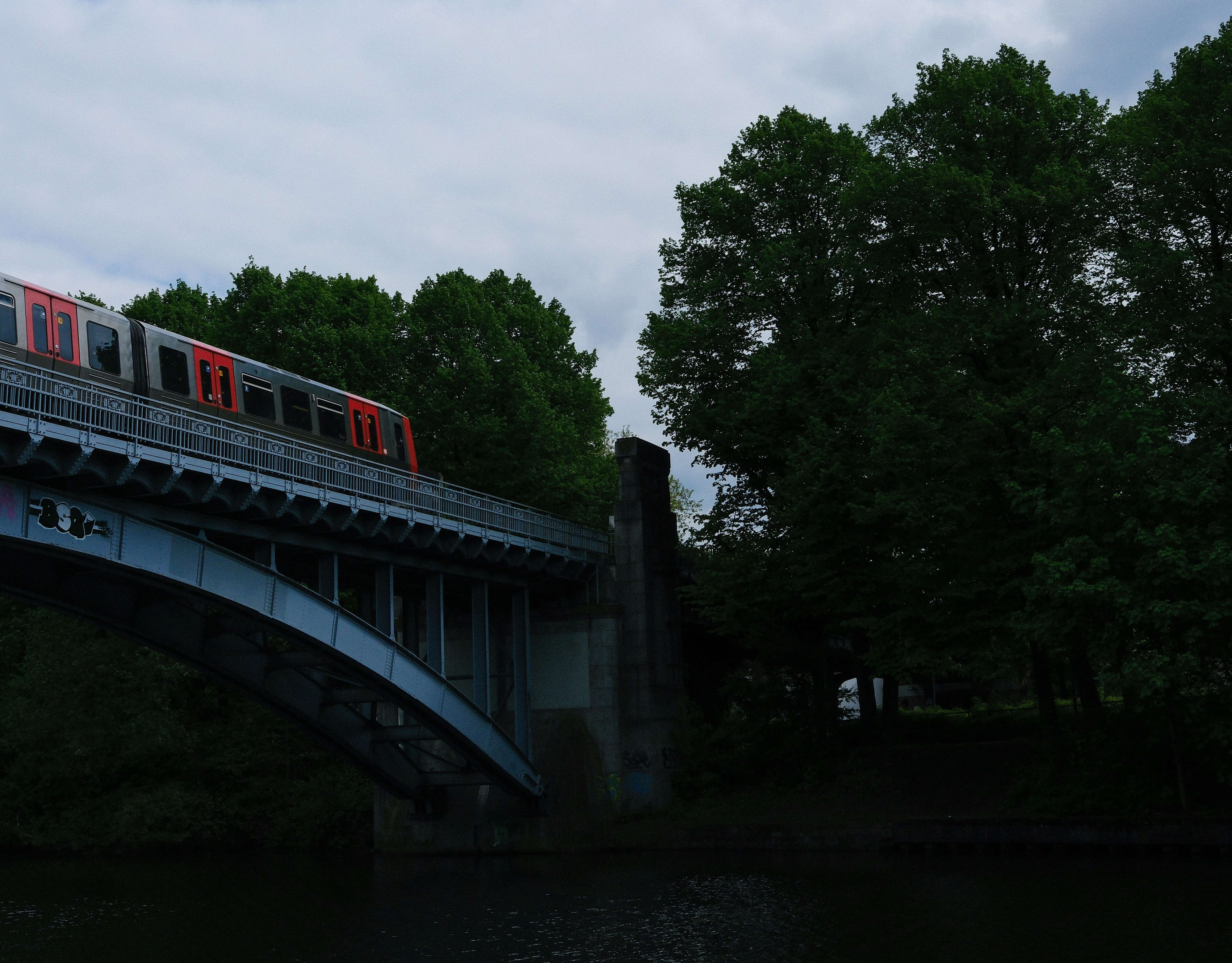 A steel-arched bridge carries a commuter train above a quiet river park