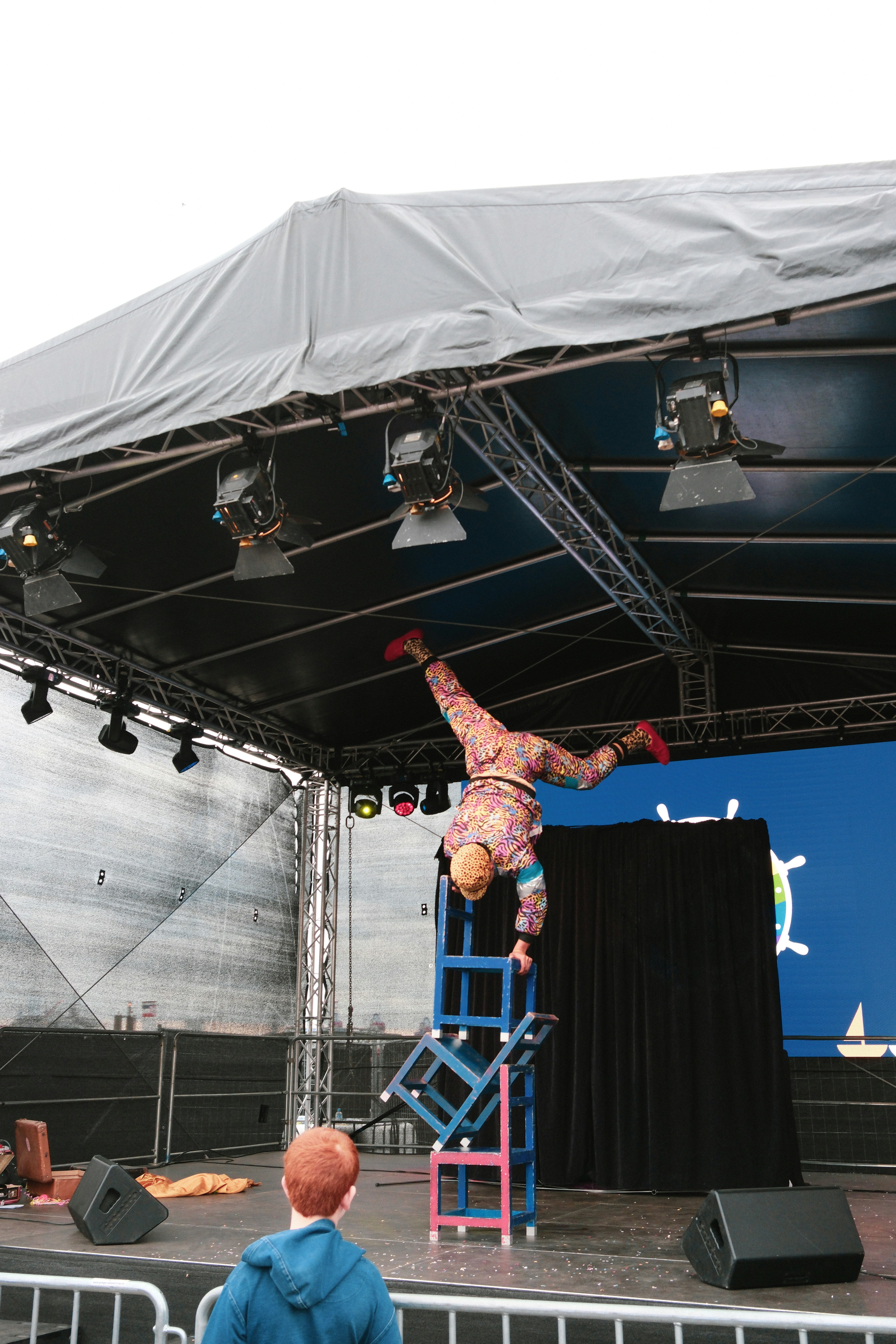 Colorful performer balances on stacked chairs during a stage performance, with an audience member in the foreground and a blue backdrop behind the act.