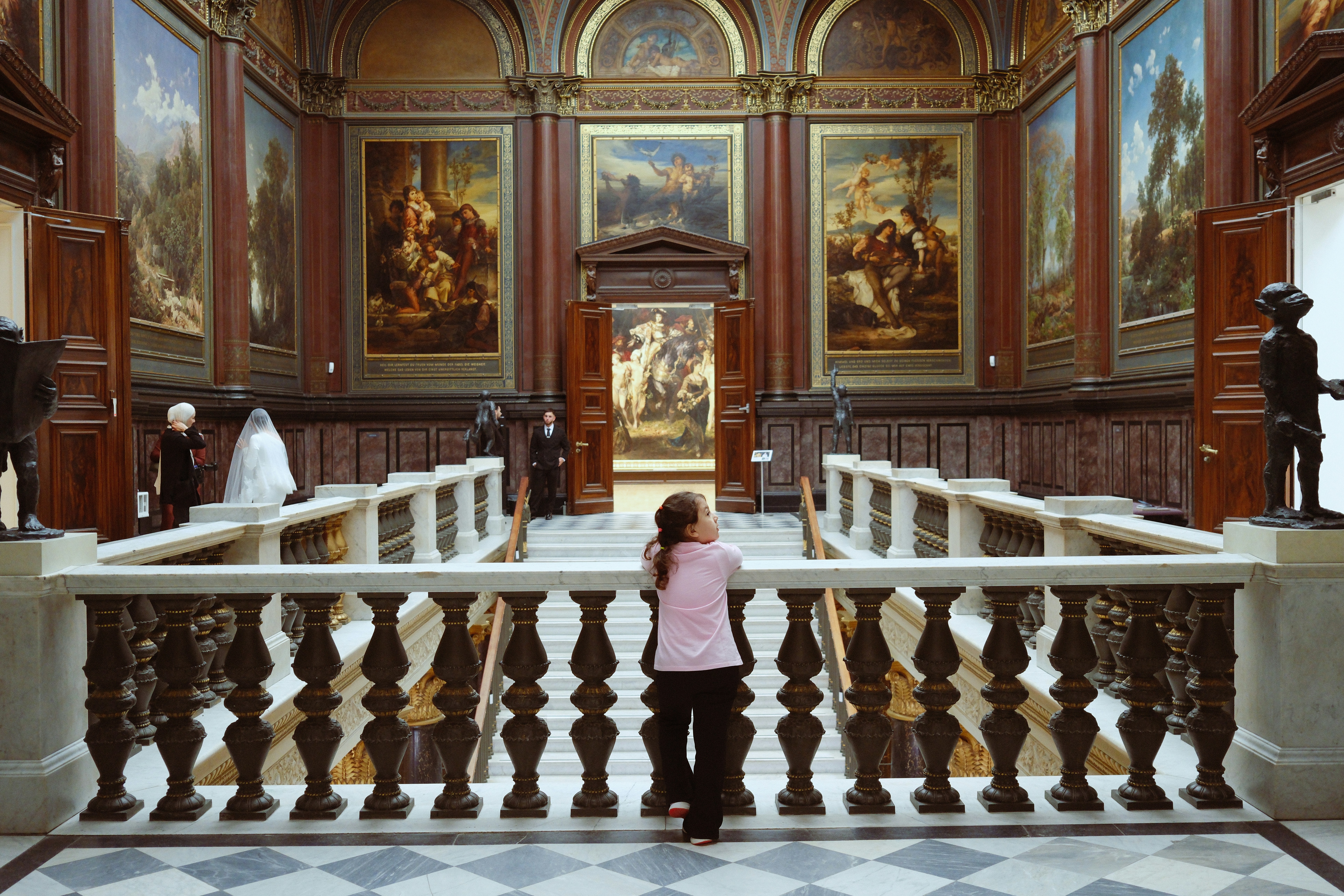 Centered in a grand gallery: a child imitating statues before a painting gallery