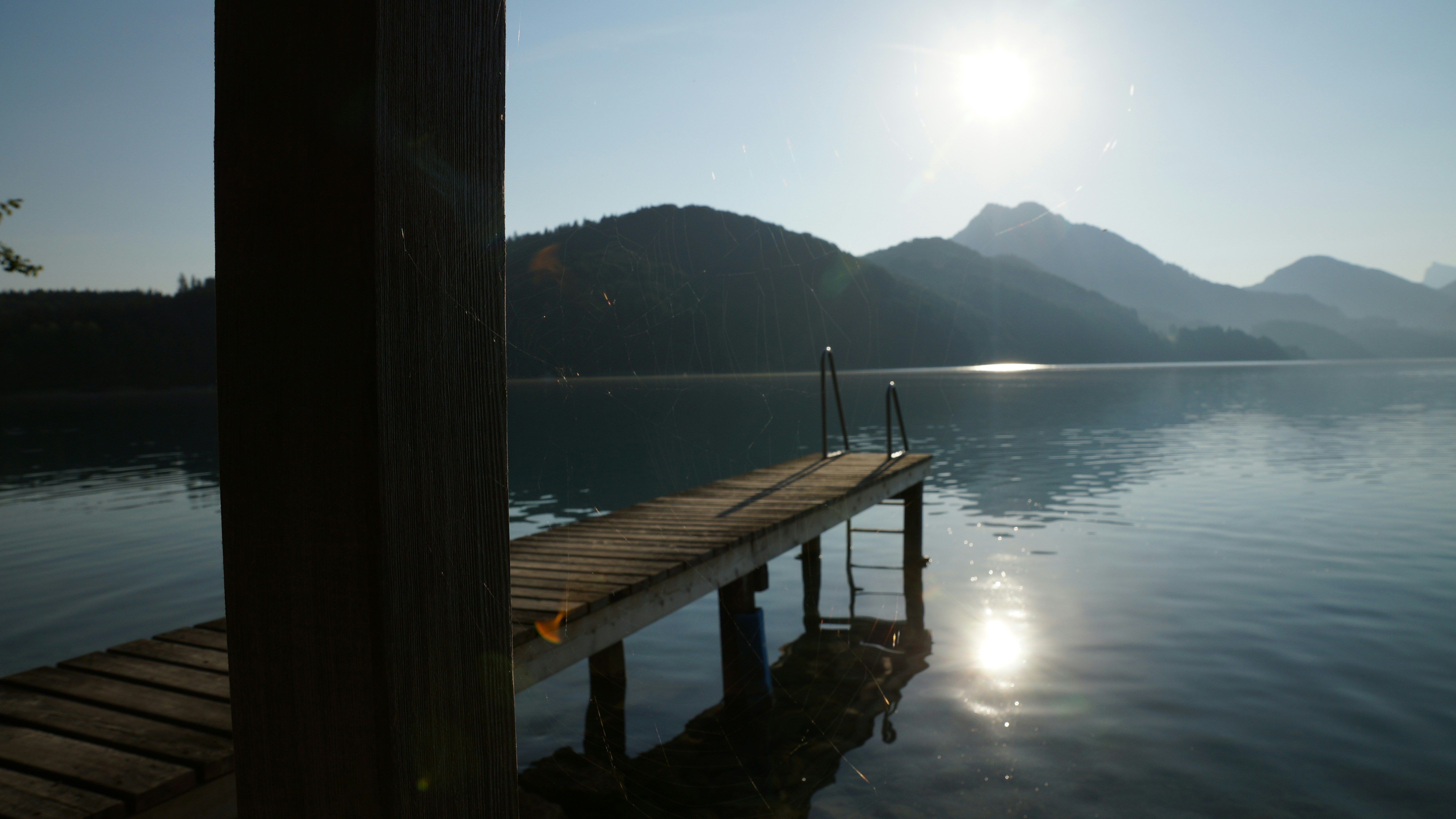 Sunlit Silence: A Wooden Pier Reaches Toward Quiet Pines and Distant Peaks