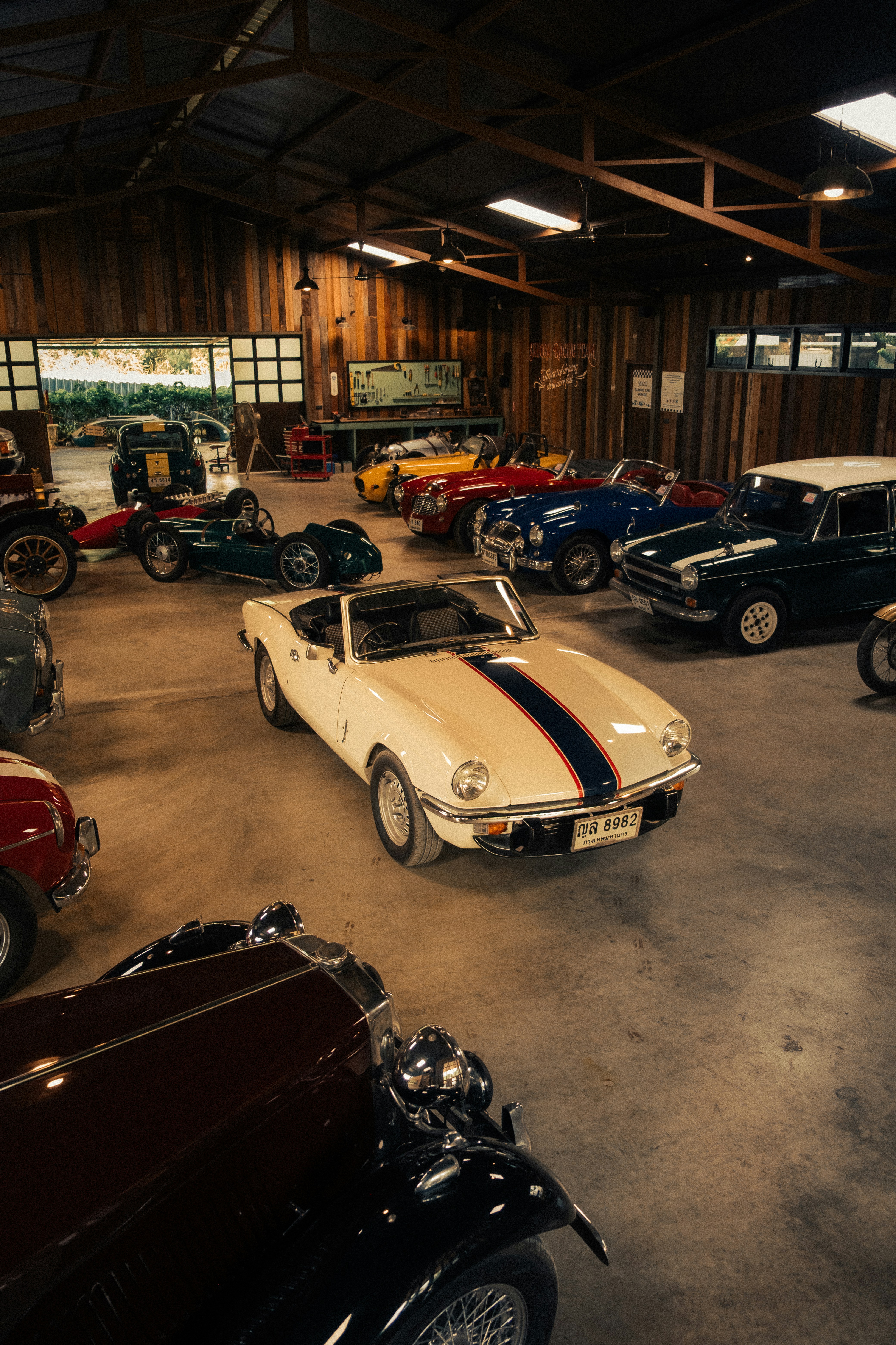 Cream sports car with blue racing stripe sits center in a rustic wood-constructed showroom filled with classic cars. A mix of convertibles and coupes line the workshop, with natural light entering from the far side.