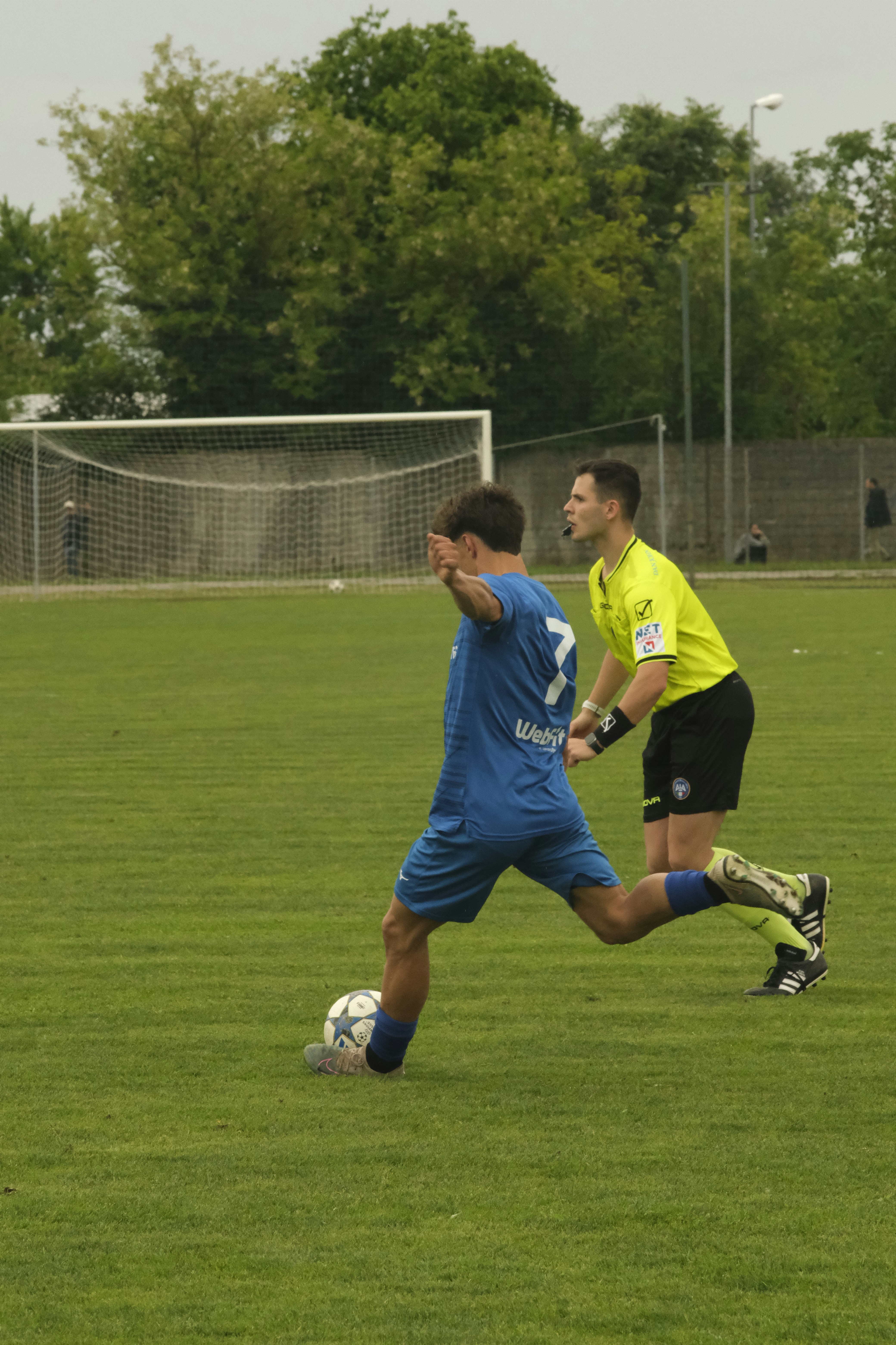 Blue striker and yellow referee chase a advancing ball on a sunlit grass pitch