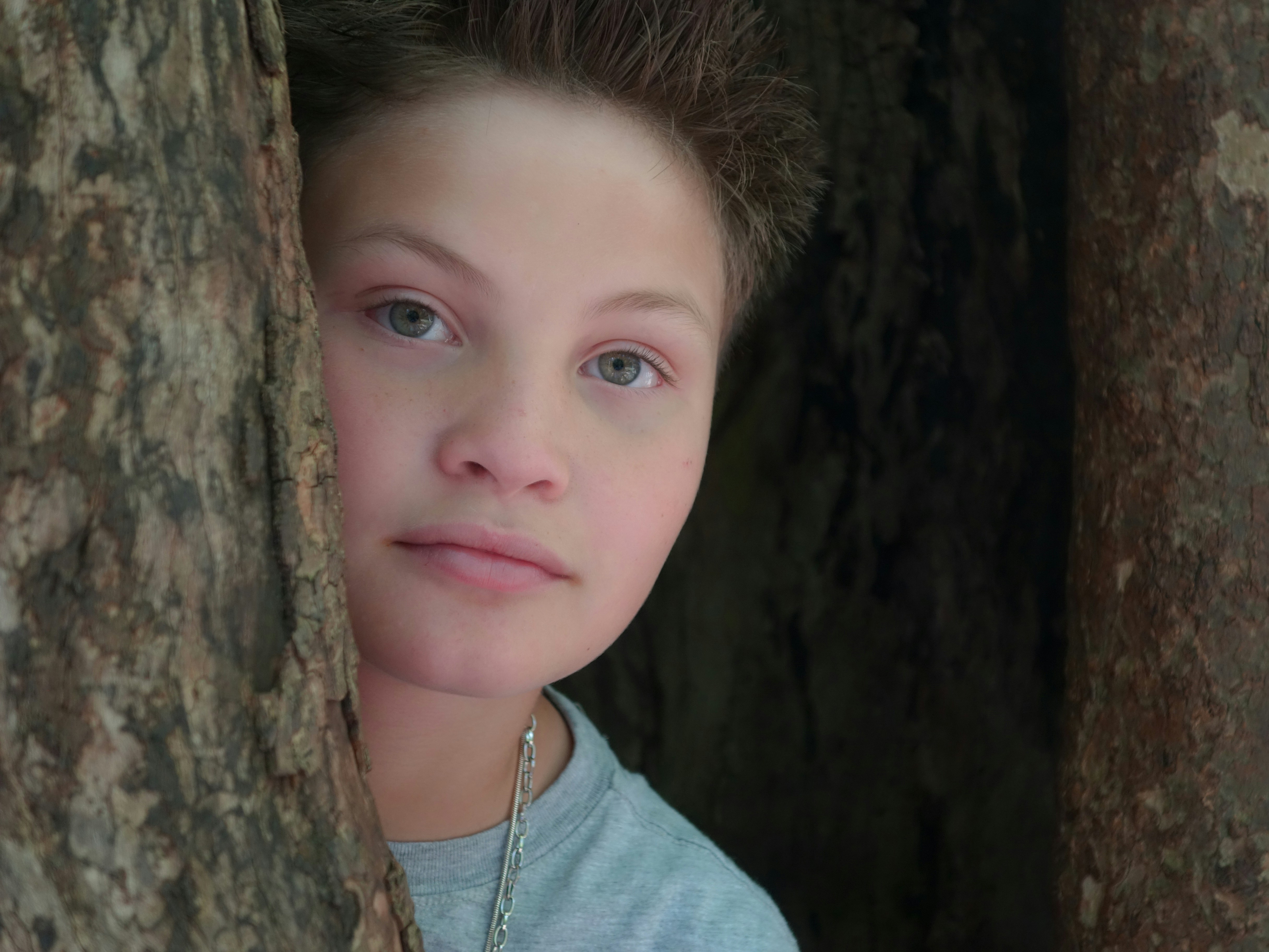Portrait of a young girl peeking from between tree trunks with soft natural light on her face; a subtle backdrop of dark forest creates a calm, intimate mood.