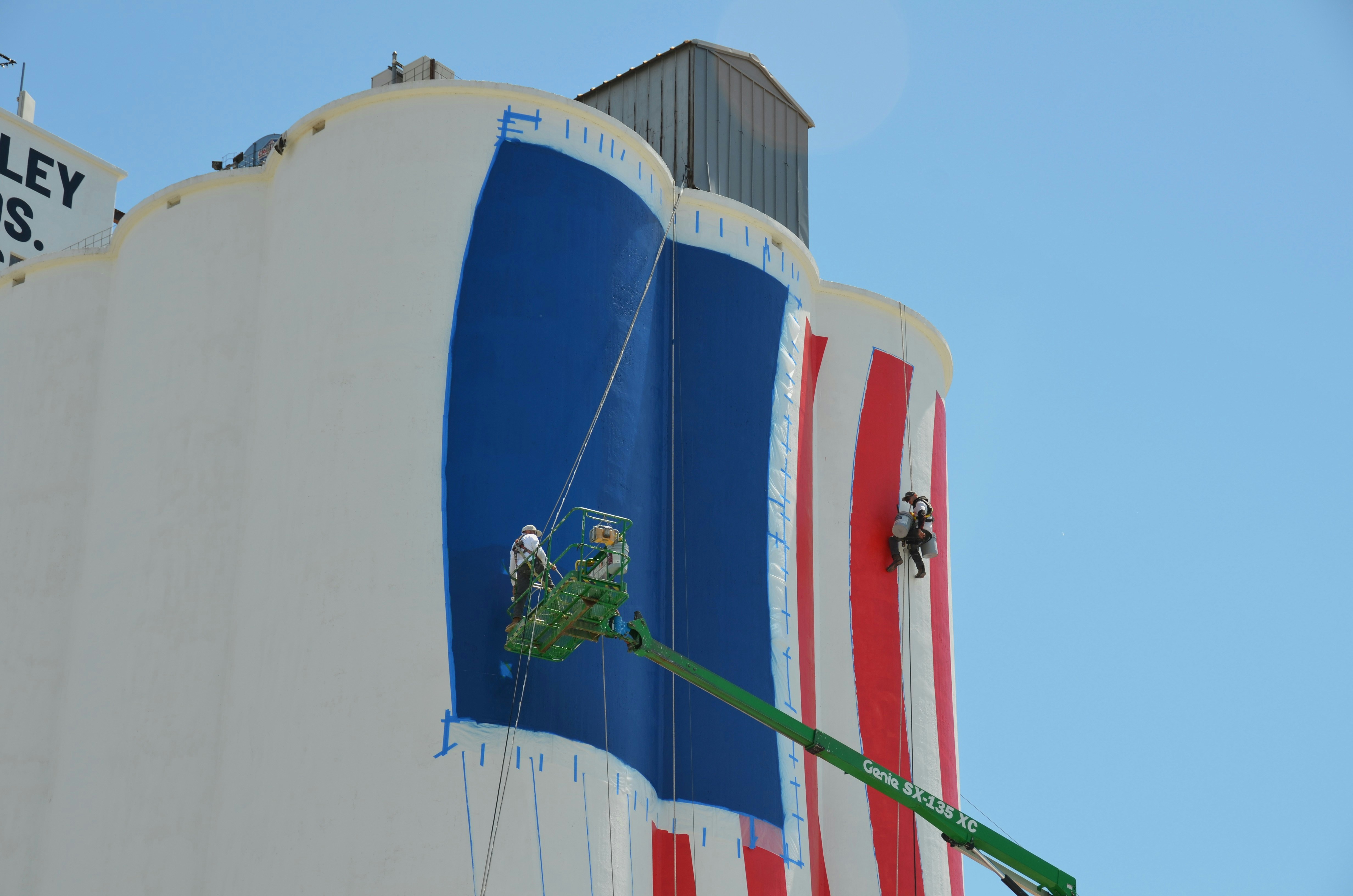 Flag-adorned silo under construction, lifted by a green boom reaching into a clear blue sky