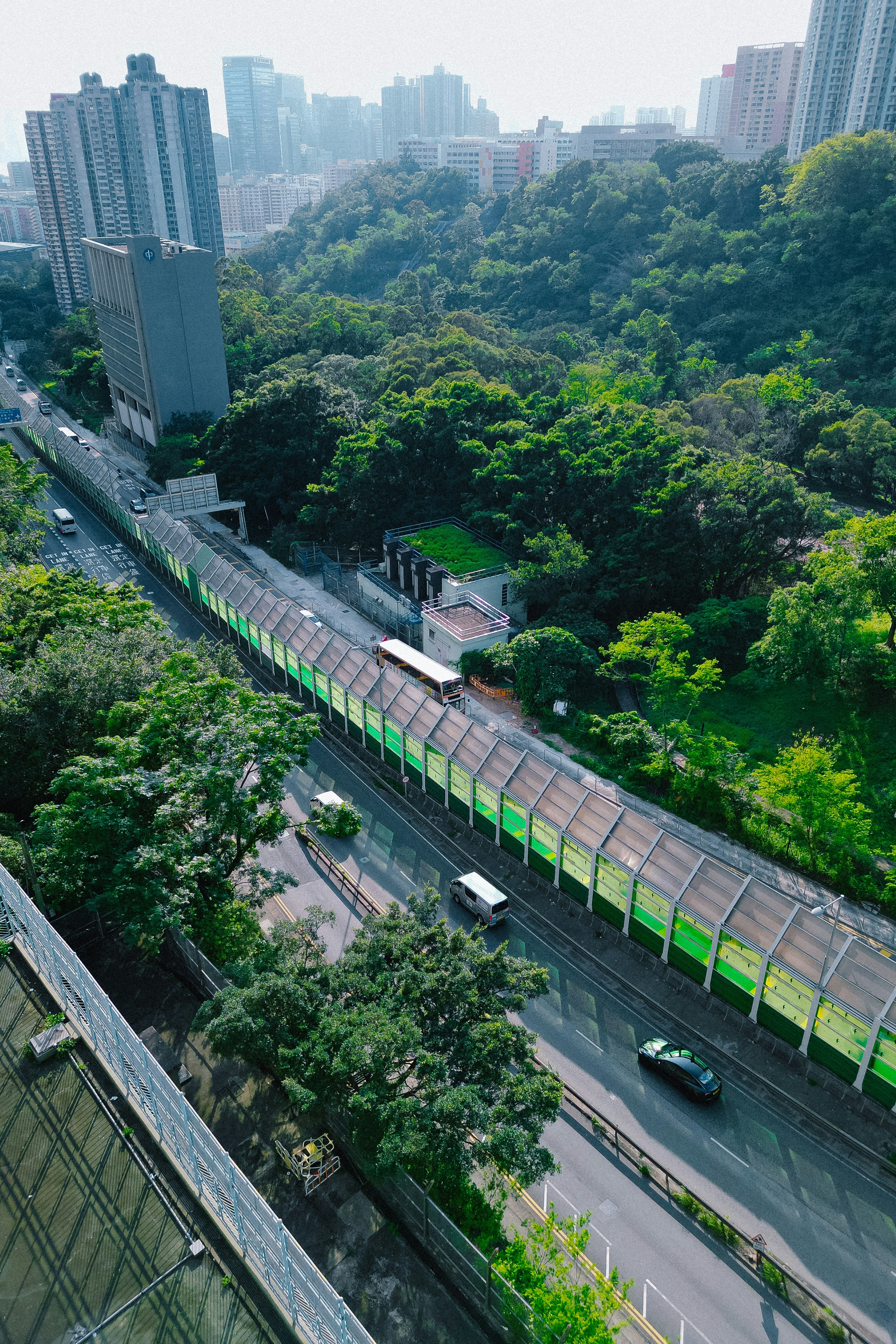 Highway with green sound barrier, lush hills, and city buildings.