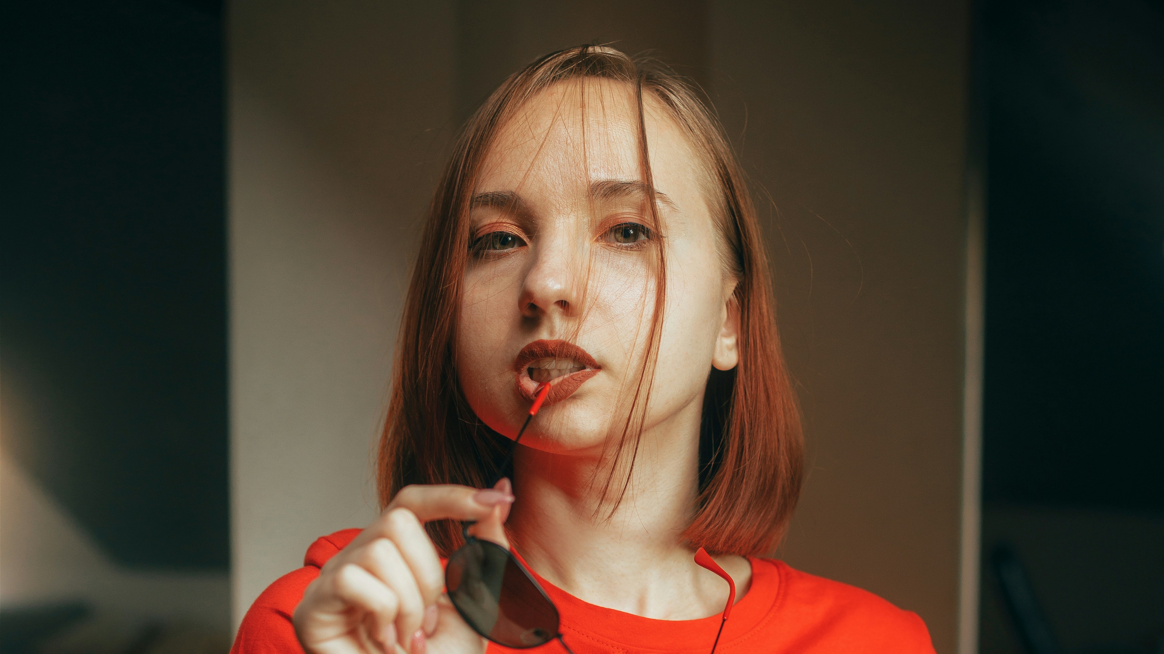 Close-up portrait of a young woman with red hair and a red top, a lipstick-stained finger near lips, against a neutral background.