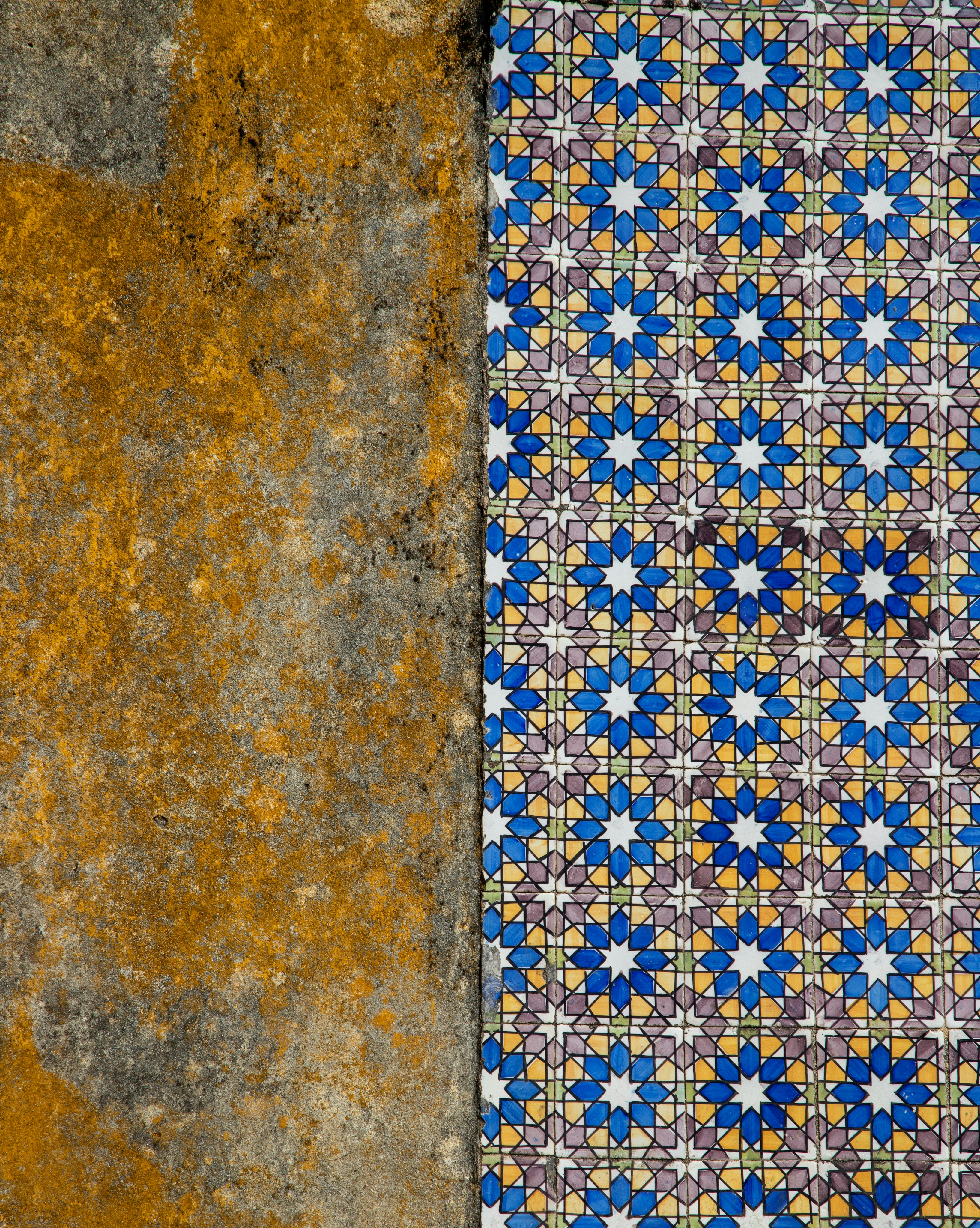 A weathered, rust-toned stone surface sits beside a vivid geometric tile pattern, creating a stark material contrast. The image highlights texture and color in a compact vertical division, suggesting everyday architectural detail. Logo: none.