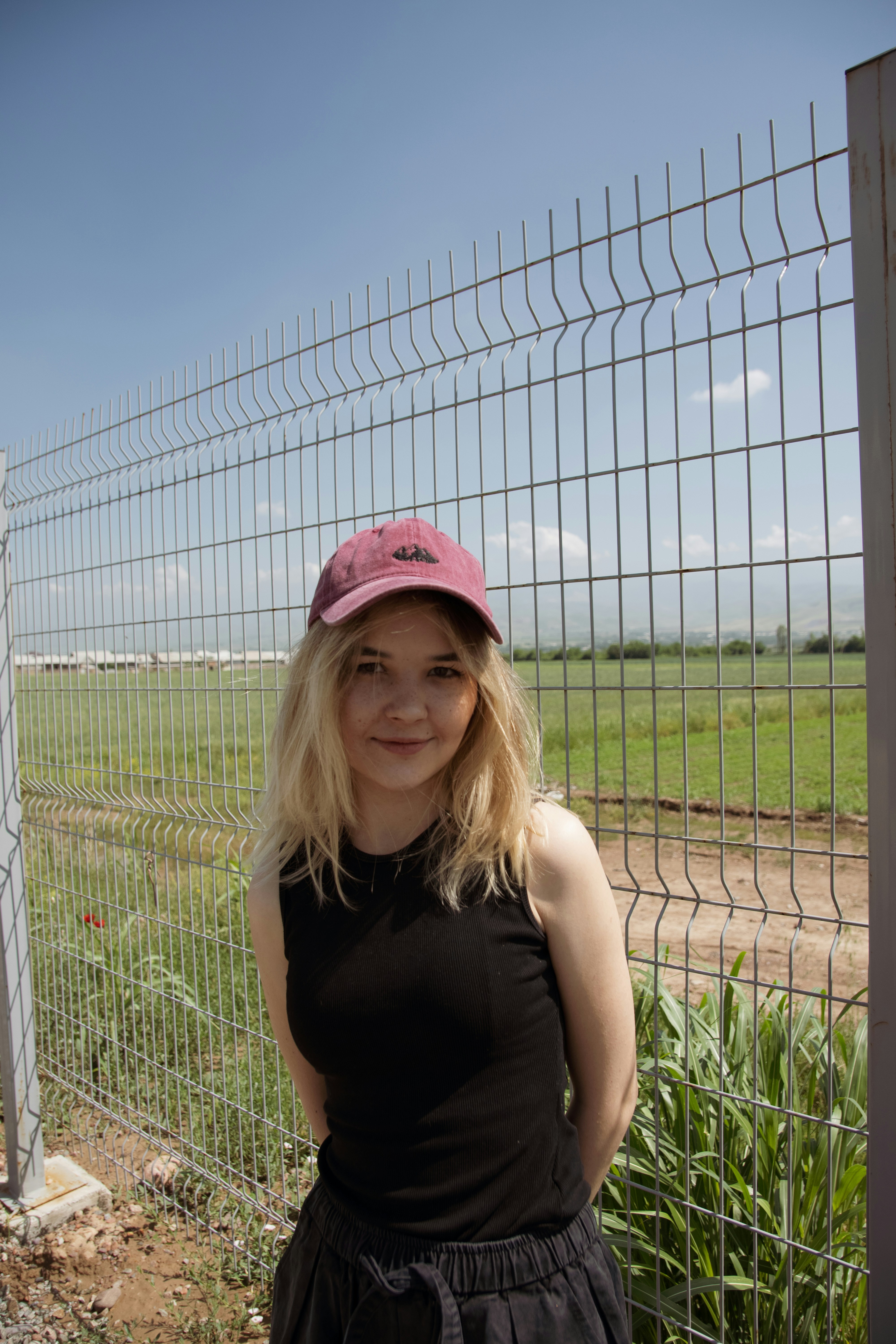 Blonde in a Pink Cap Stands by a Grid Fence Under Wide Skies