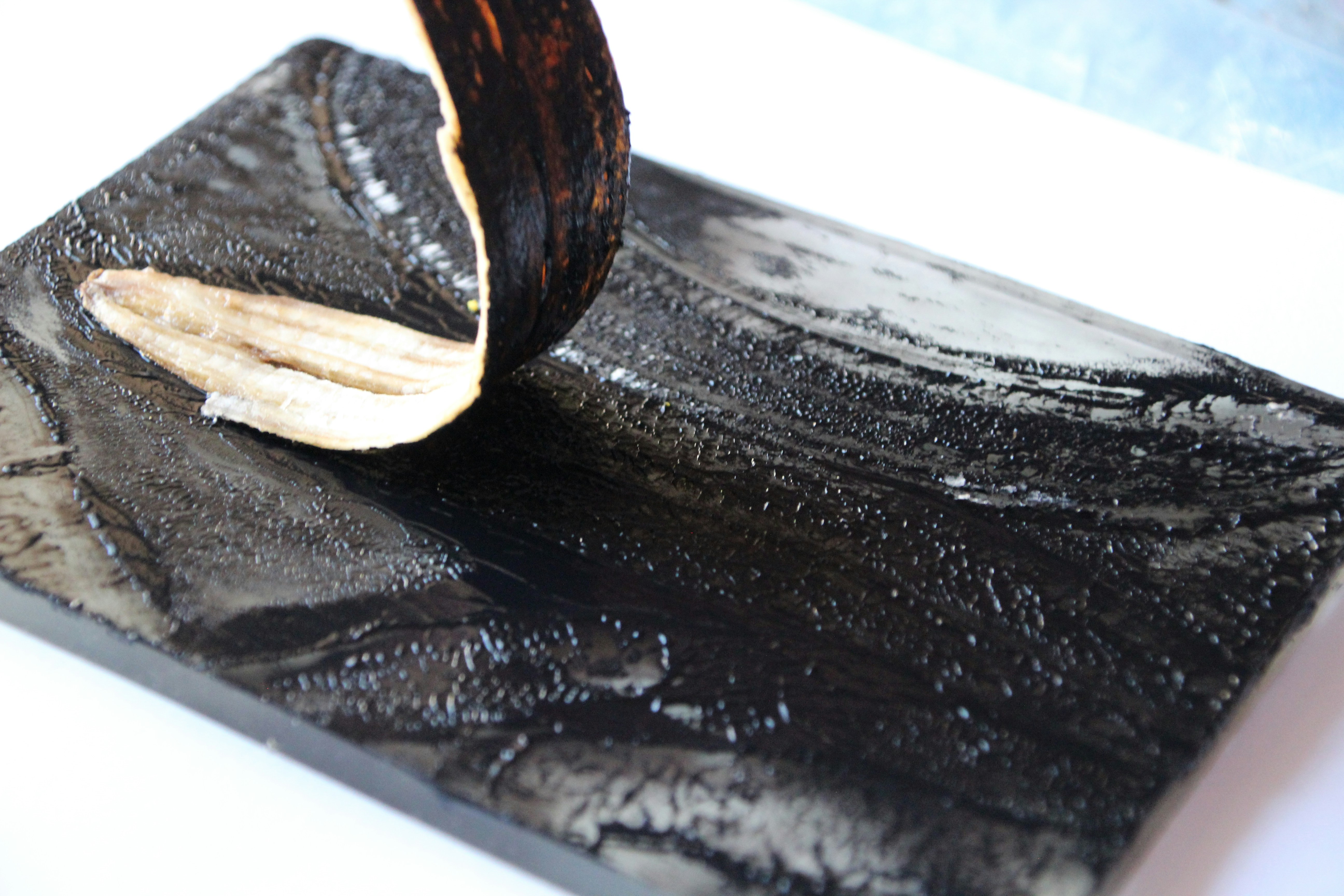 Close-up of a black glossy sheet with a curled brown edge peeling away, revealing a pale interior; the scene emphasizes surface texture and sheen against a white, softly blurred background.