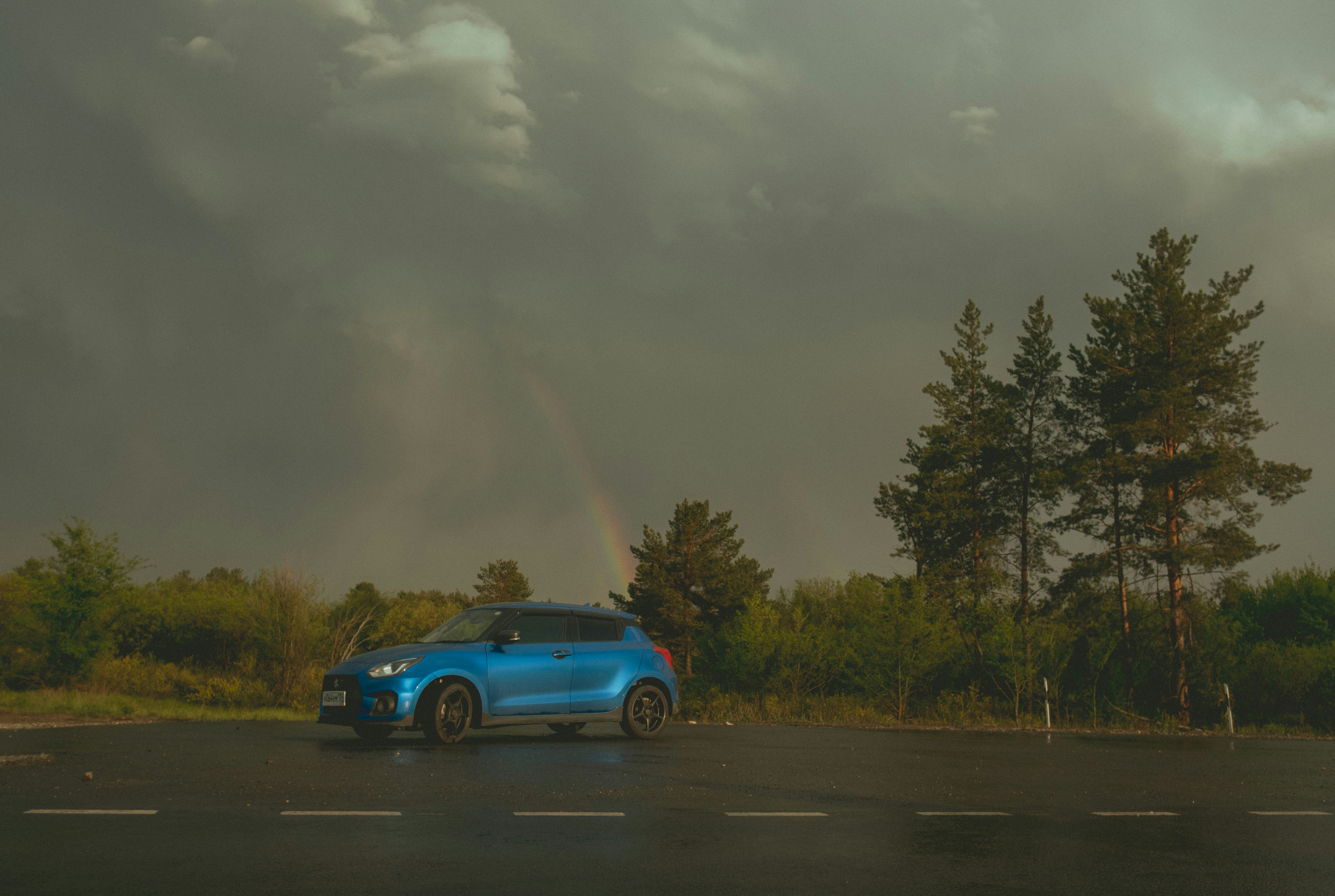 Blue hatchback under a rainbow in a rain-washed lot