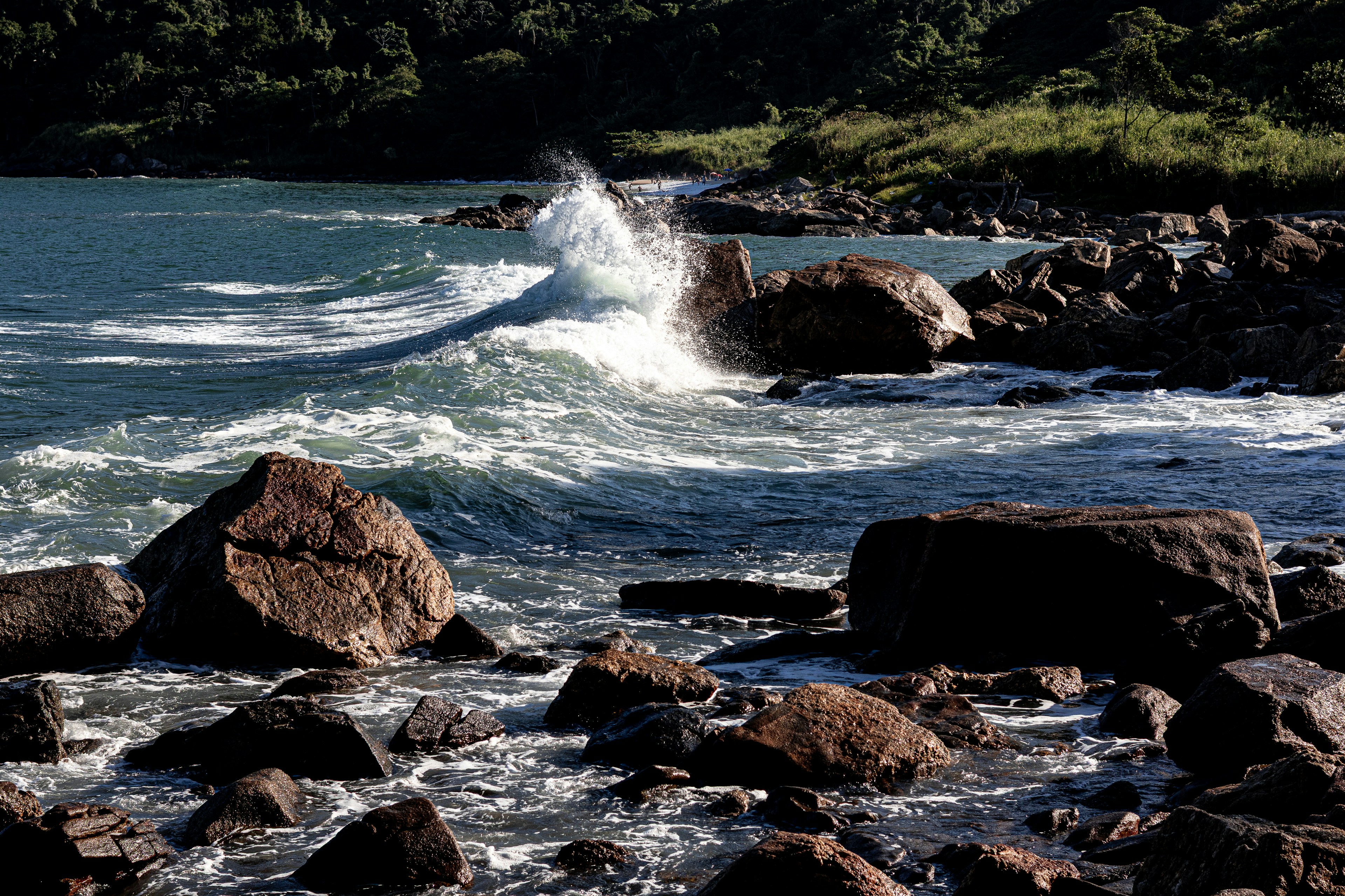Rugged rocky shoreline with a breaking wave and spray, sunlight highlighting textured surfaces and dark rocks along a blue-green sea. A natural landscape shot without people, emphasizing motion and contrast.