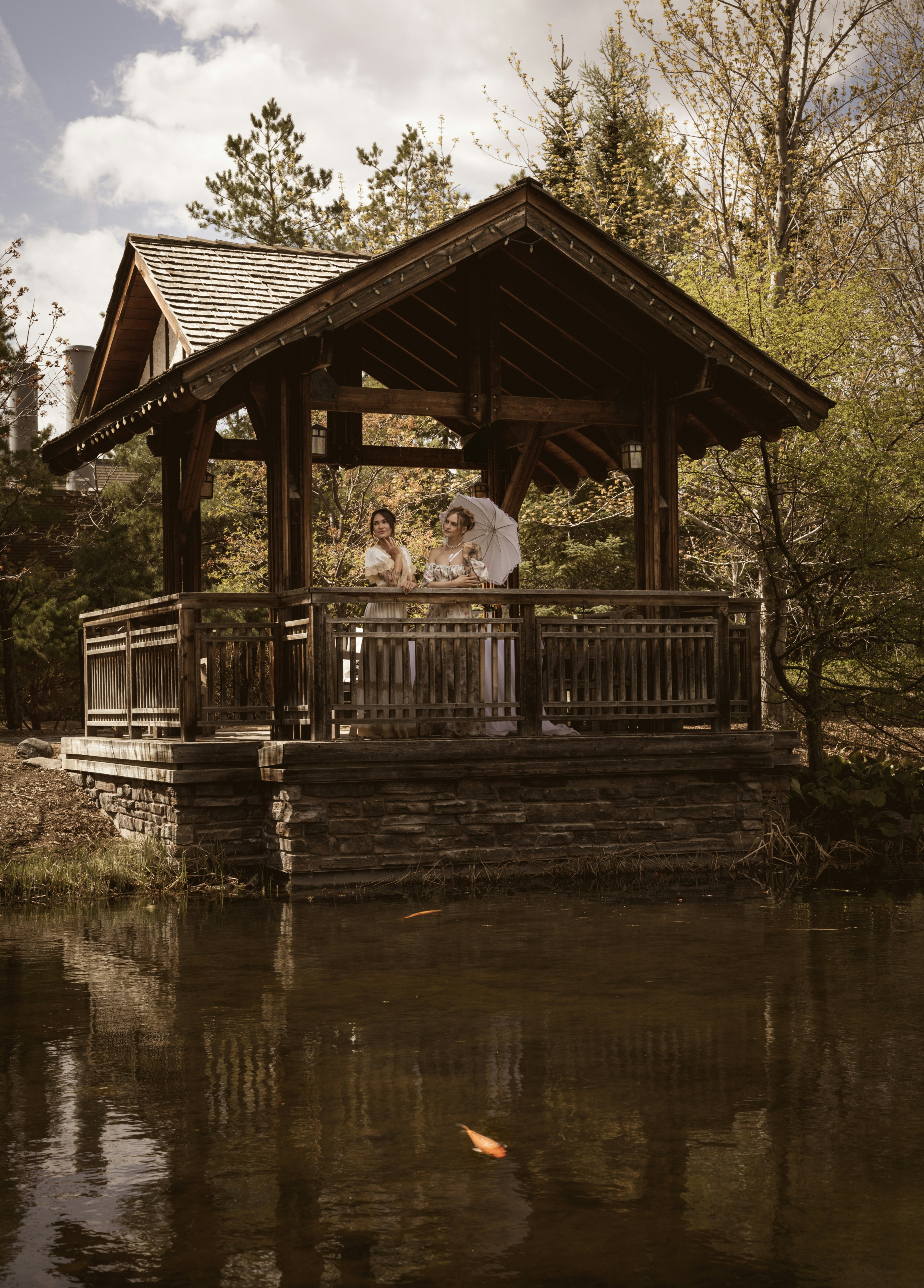 Two women in dresses under a wooden gazebo by water.