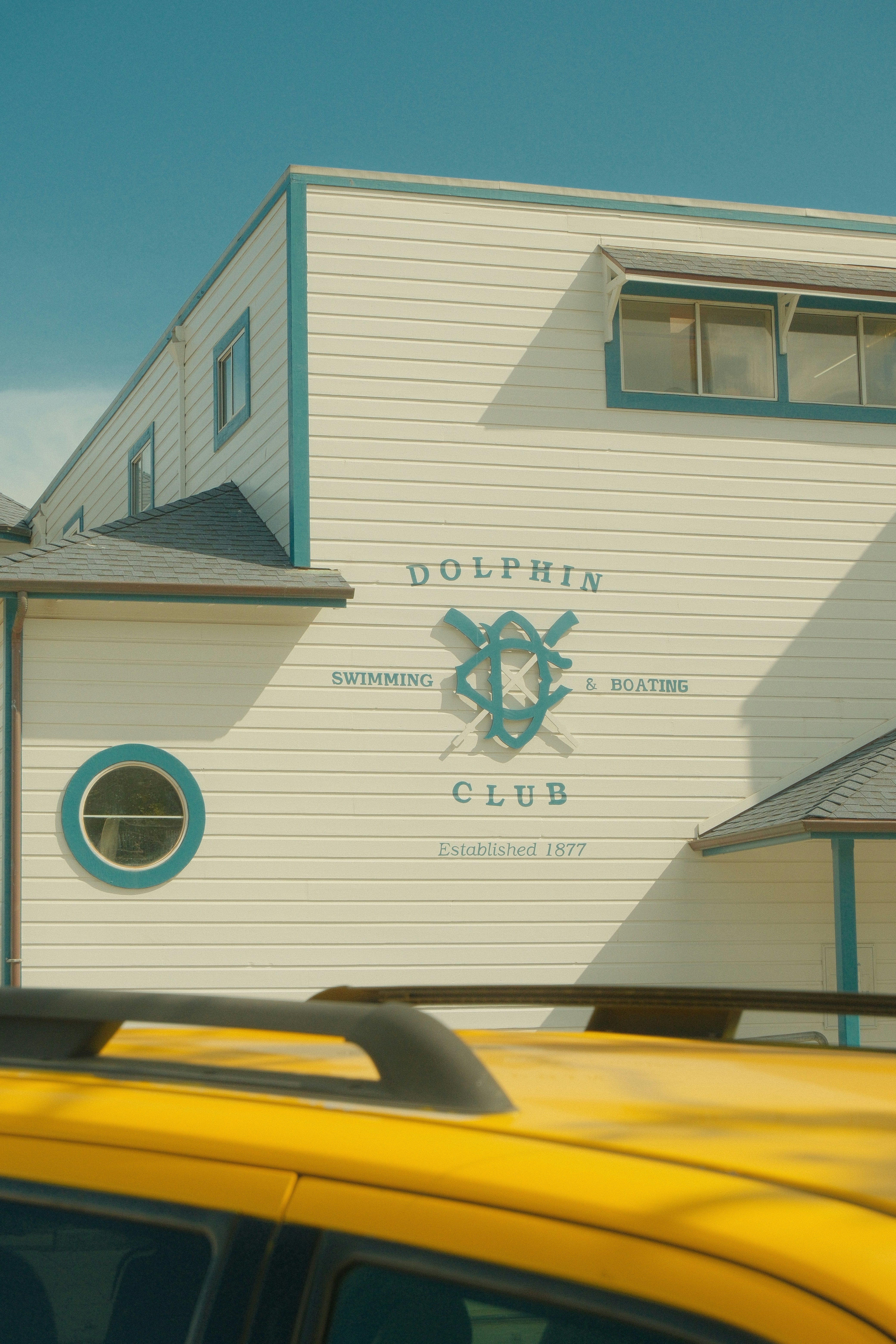 Dolphin Club Signage on Sunlit White Building with Bright Yellow Car Roof
