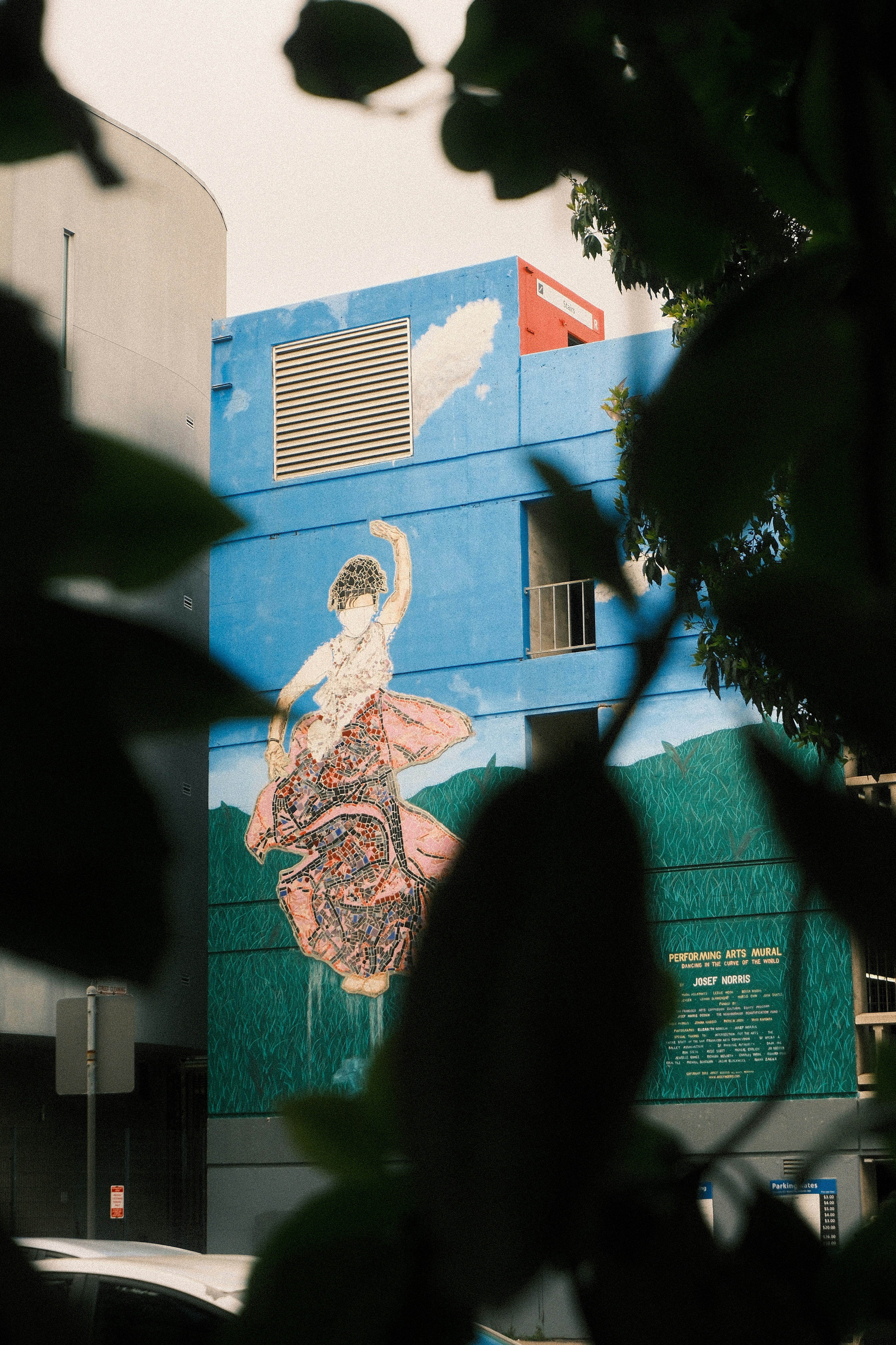 Dancer on a mural: blue building framed by leafy shadows