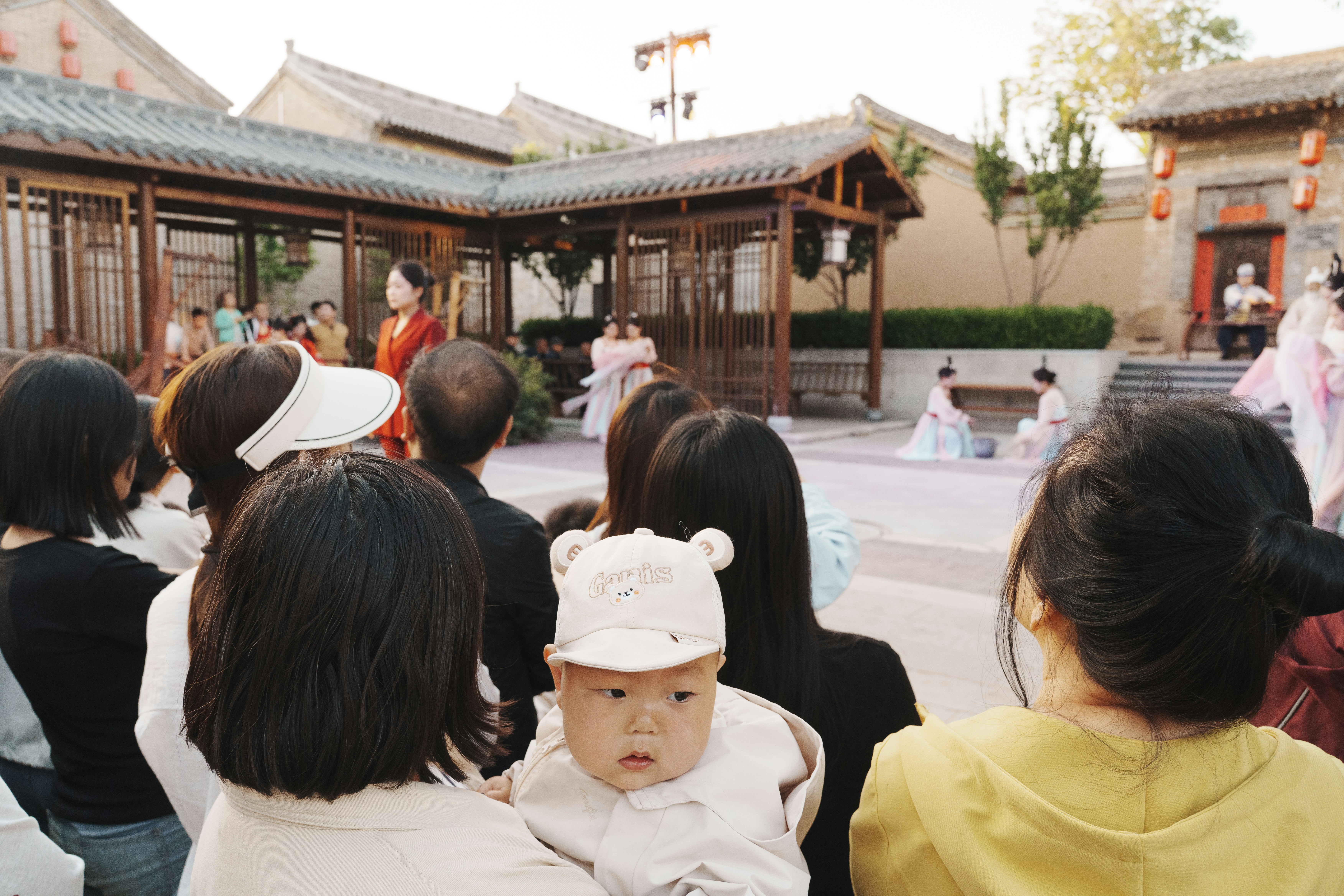Child in a white cap overlooked by a crowd at a traditional courtyard gathering