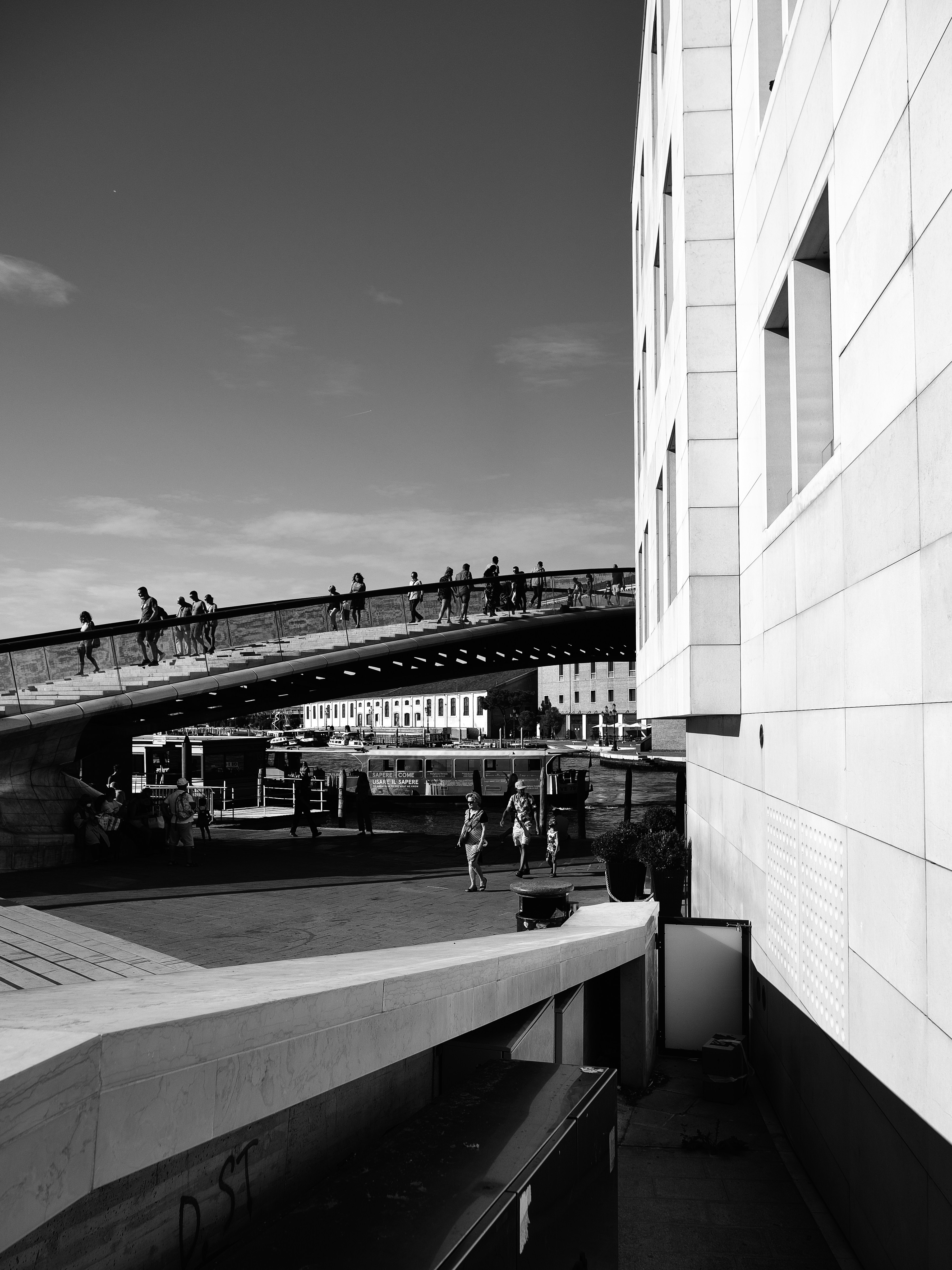 Black-and-white urban scene showing a modern building on the right, a pedestrian bridge with people crossing above a plaza below.