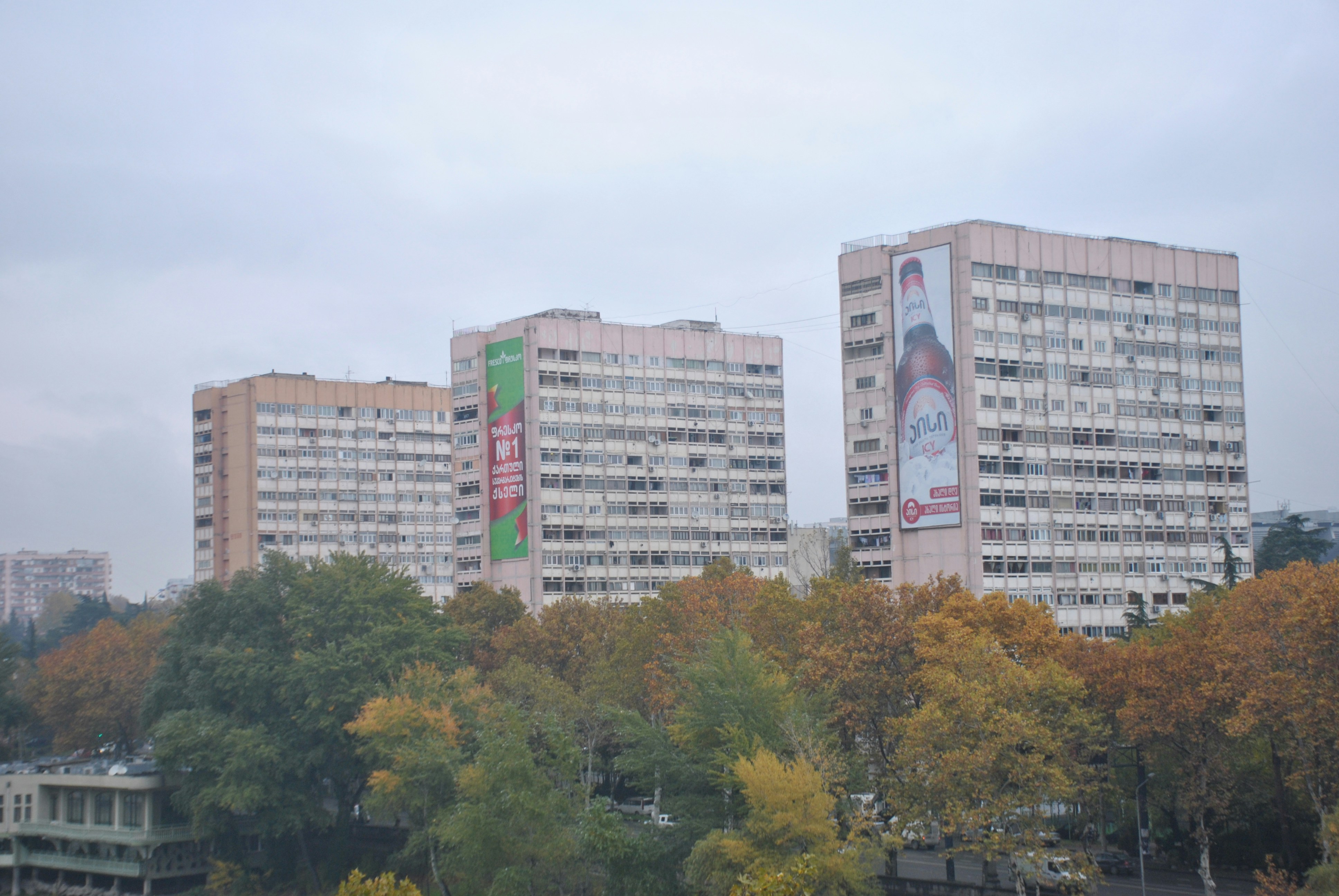 Autumn trees line the lower third of the frame with two tall apartment blocks dominating the middle, banners visible on the facades. A soft, overcast sky completes the urban scene.