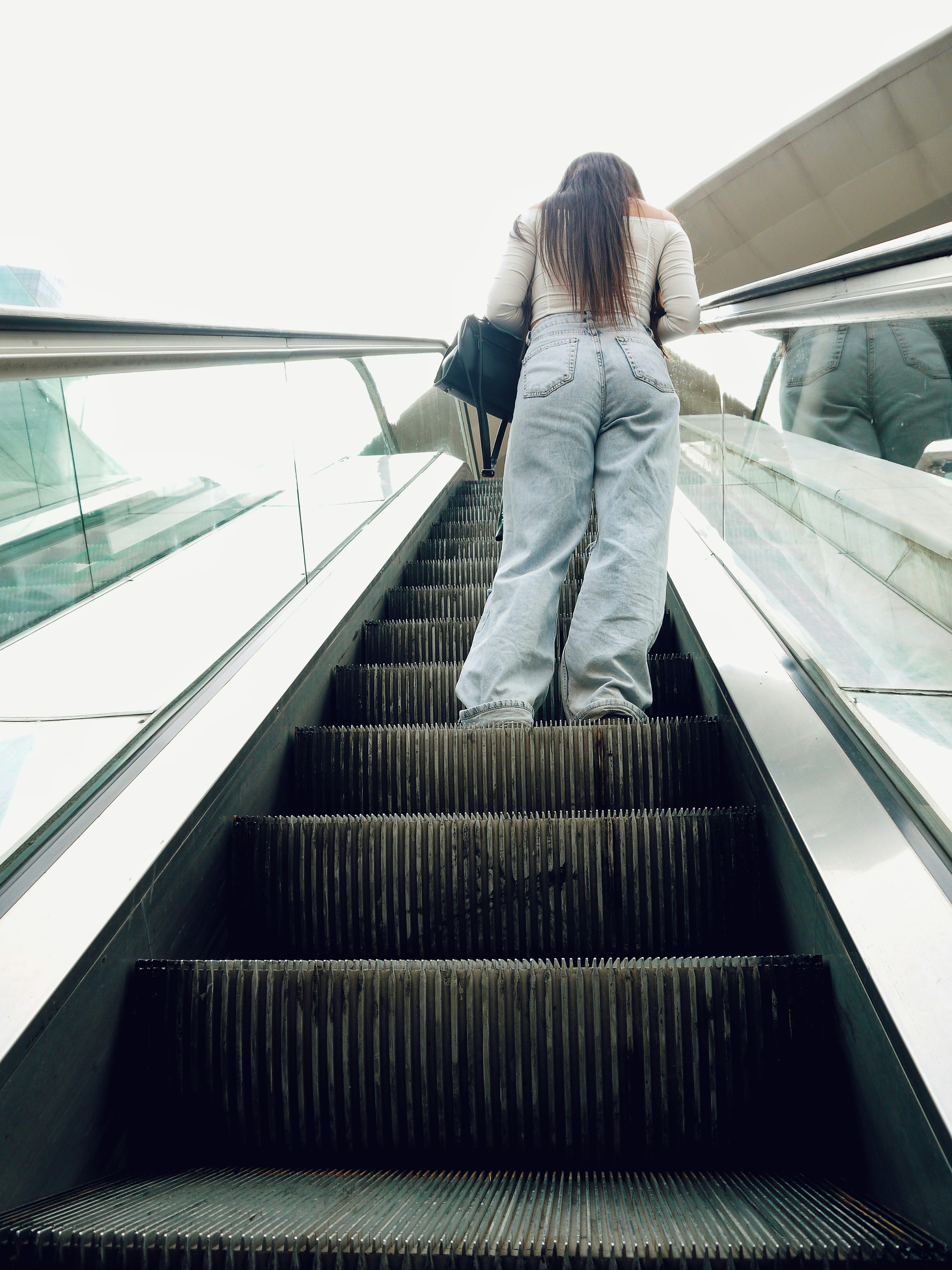 A person ascends an outdoor escalator viewed from below, jeans and a light top, carrying a bag; the scene emphasizes ascent and urban architecture with glass sides.