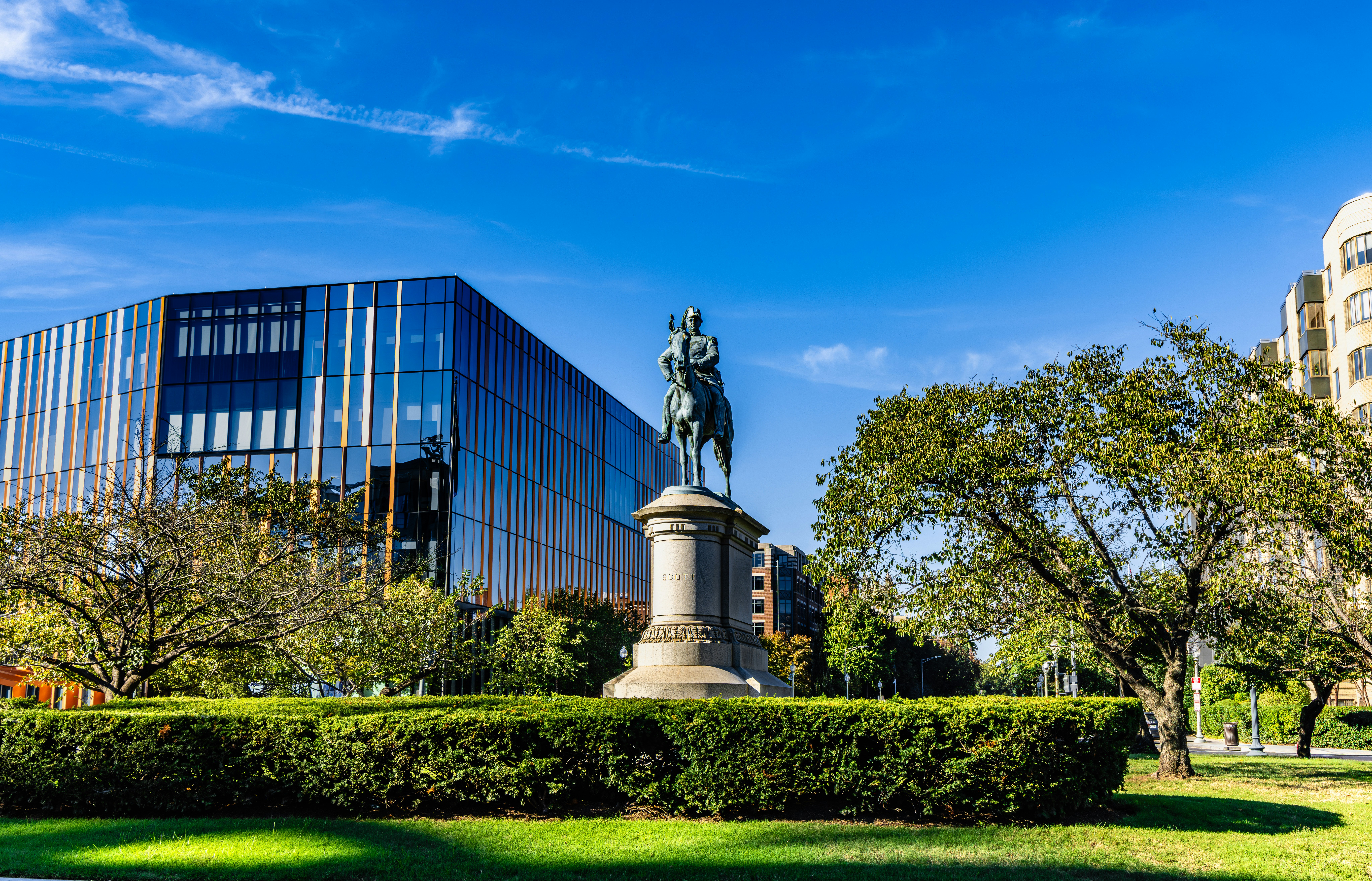 Statue in a sunlit civic plaza framed by modern glass and leafy trees