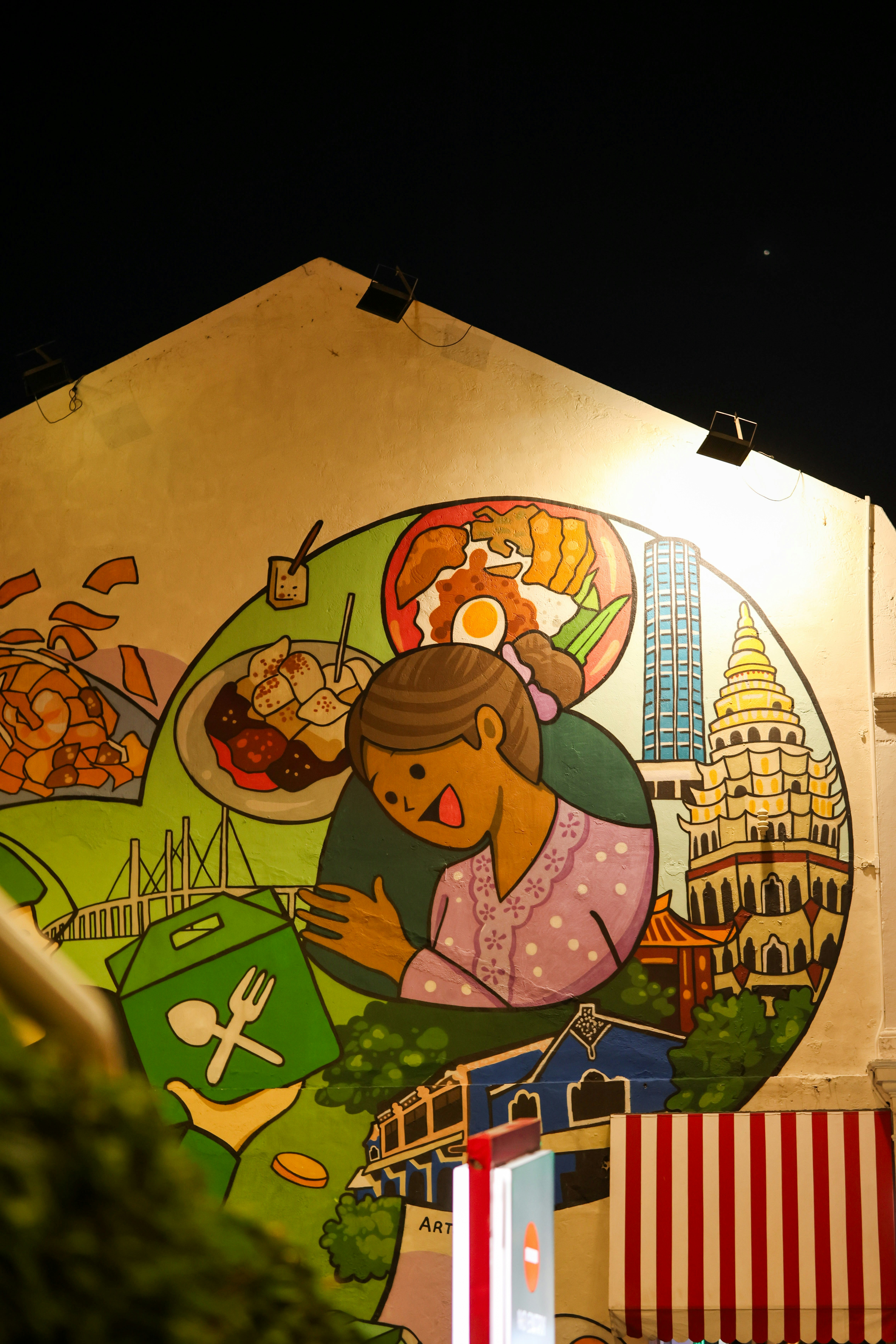 Colorful mural featuring a seated figure with hands folded, surrounded by food, architecture, and cityscape elements under warm night lighting.