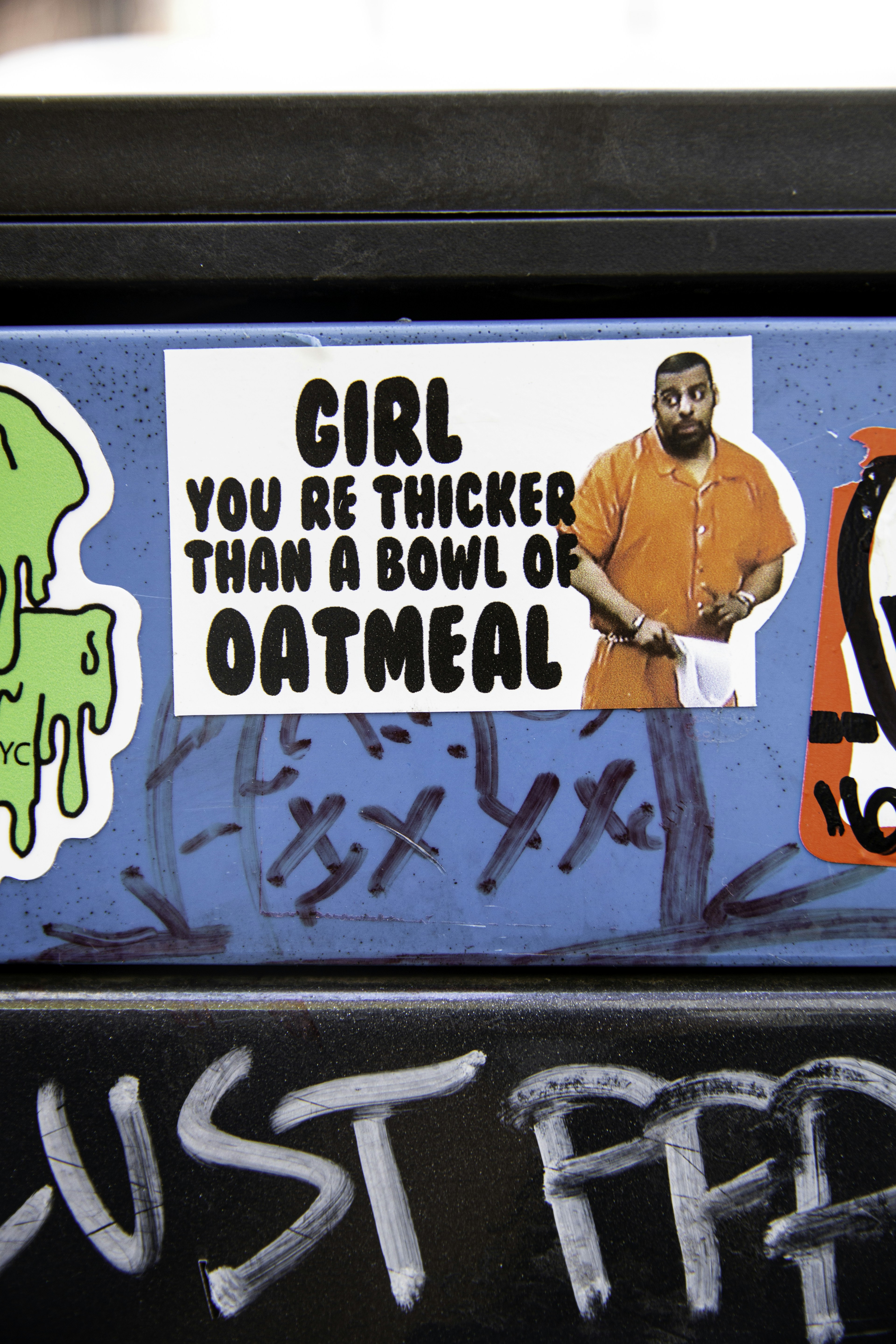 A humorous sticker featuring a man in an orange jumpsuit reads: “GIRL YOU'RE THICKER THAN A BOWL OF OATMEAL.” Surrounded by graffiti and layered stickers, the message delivers a playful street joke through collage and public humor.