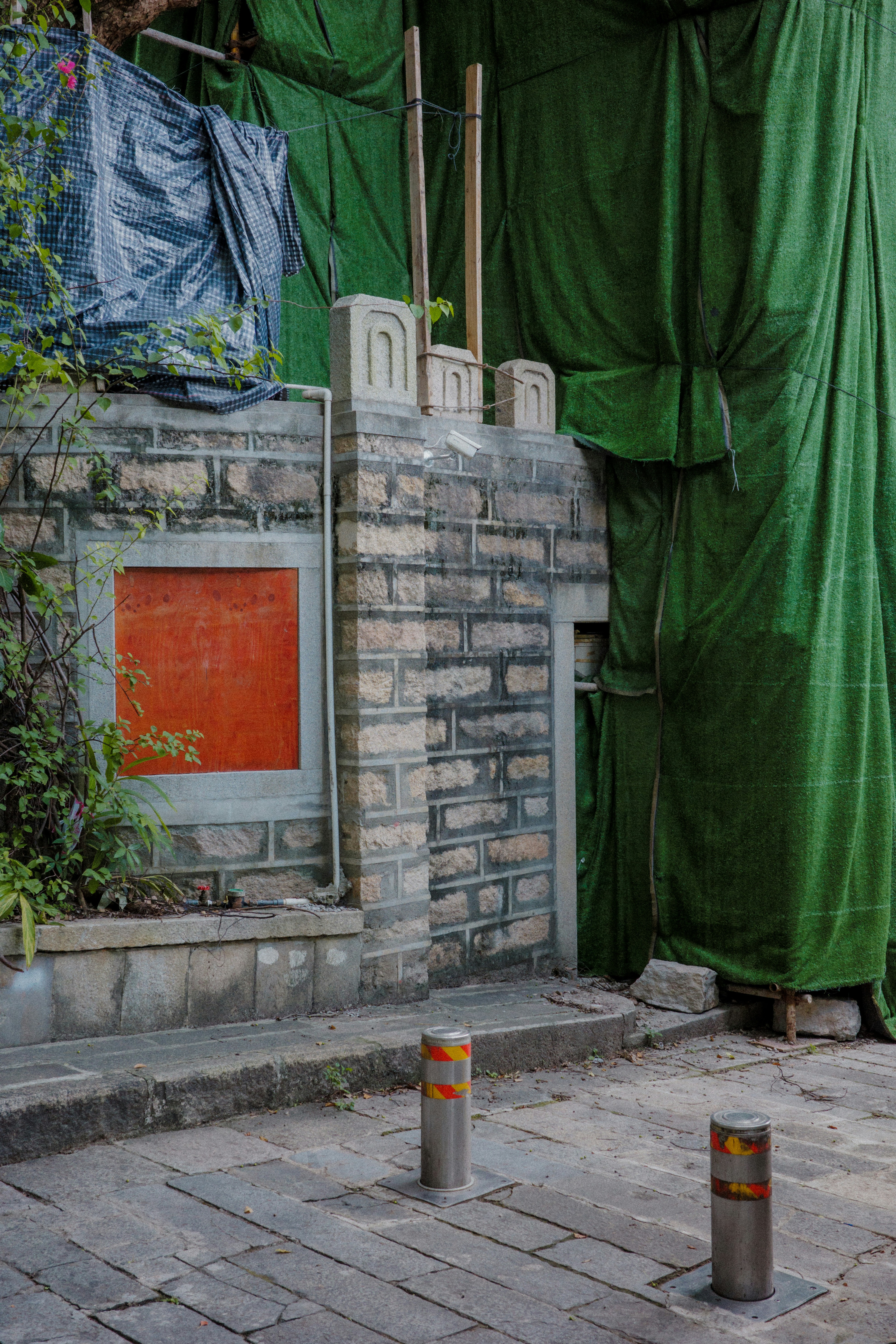 Quiet Corner of a Construction Wall: Green Tarps, Brickwork, and Steel Bollards