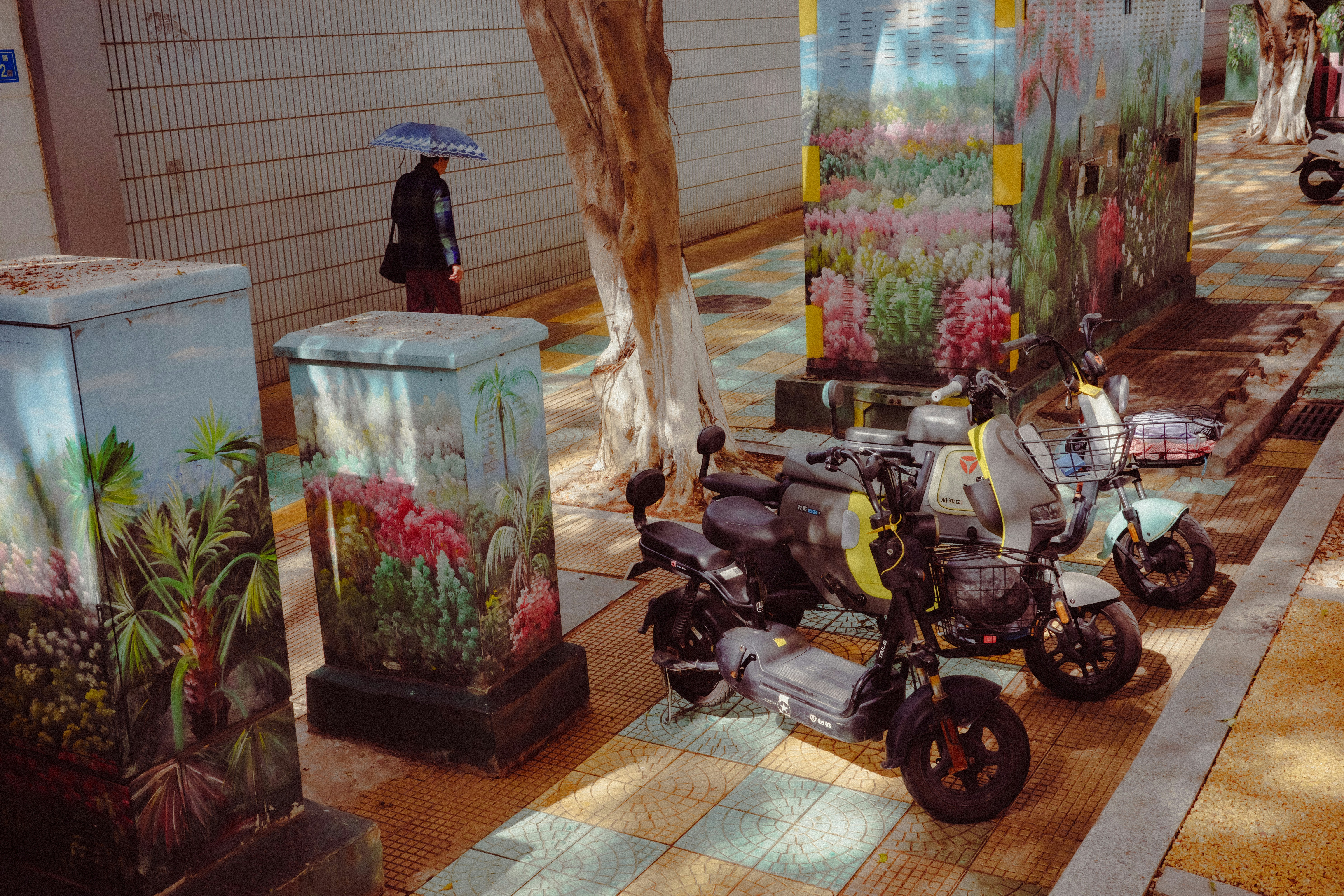 Person with umbrella walks past decorated utility boxes and scooters.