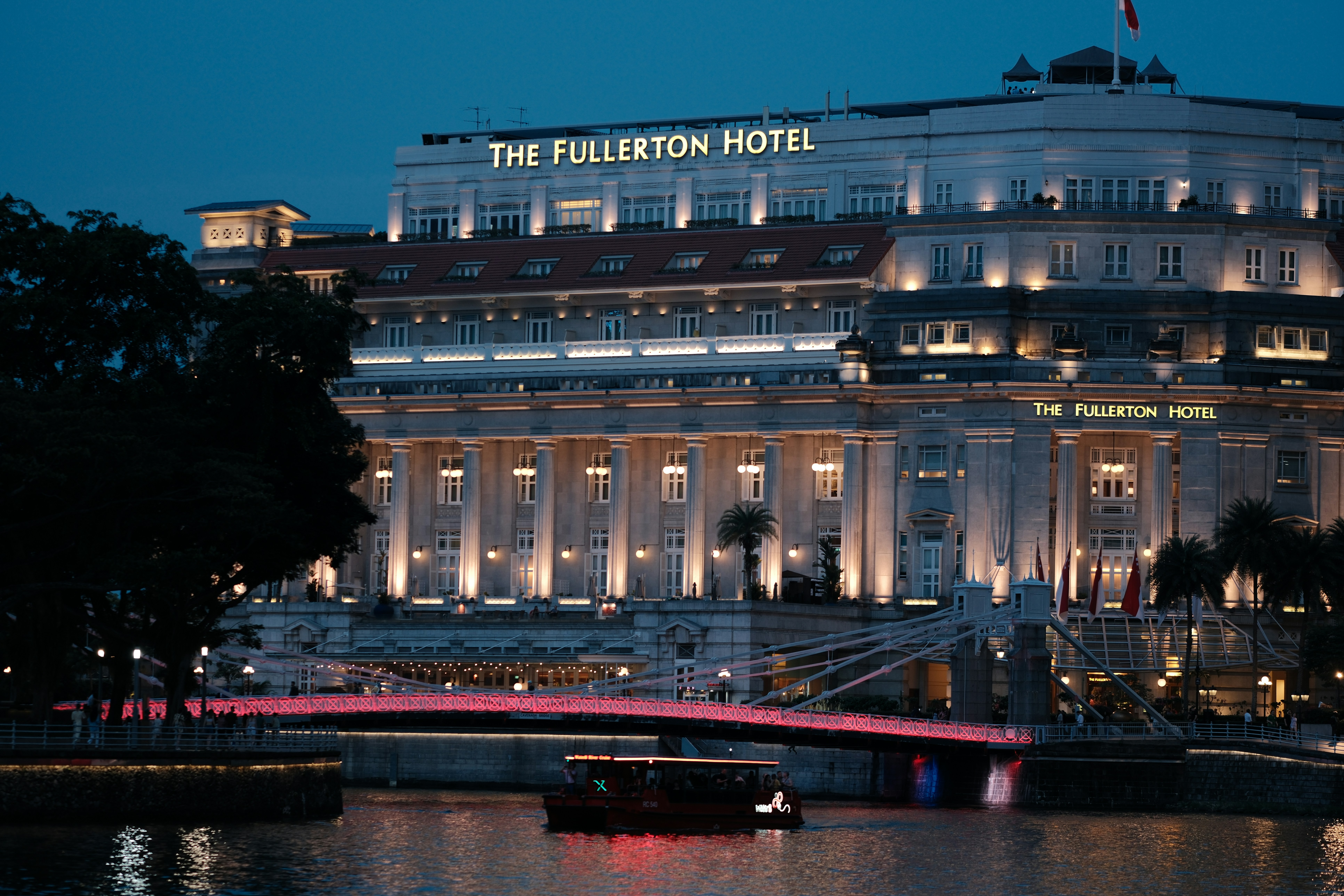 Night scene of The Fullerton Hotel illuminated along the waterfront; boat traffic and glowing reflections create a peaceful urban panorama.