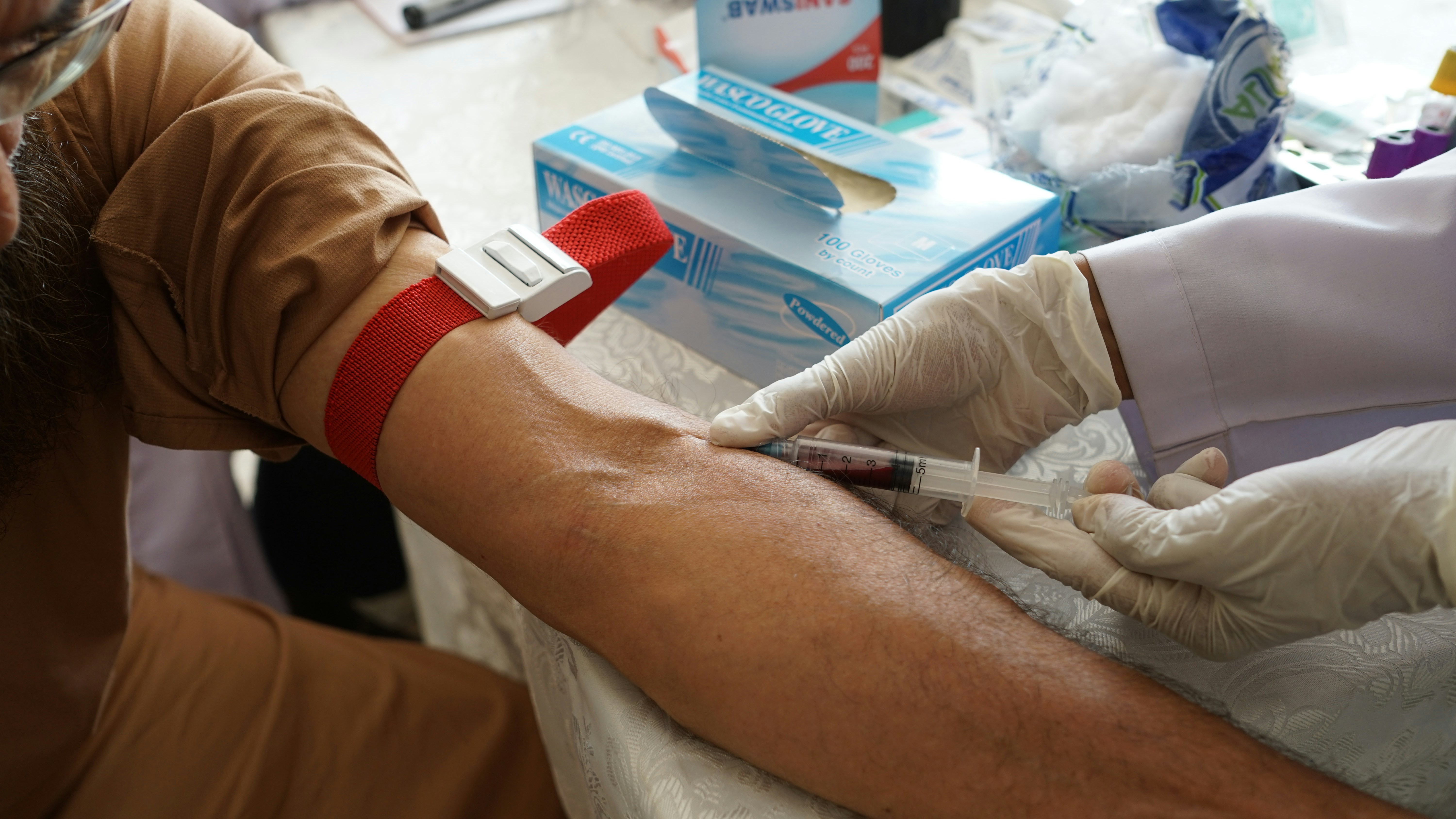 Medical procedure scene showing a gloved clinician administering an injection to a patient’s forearm with medical supplies in the background.