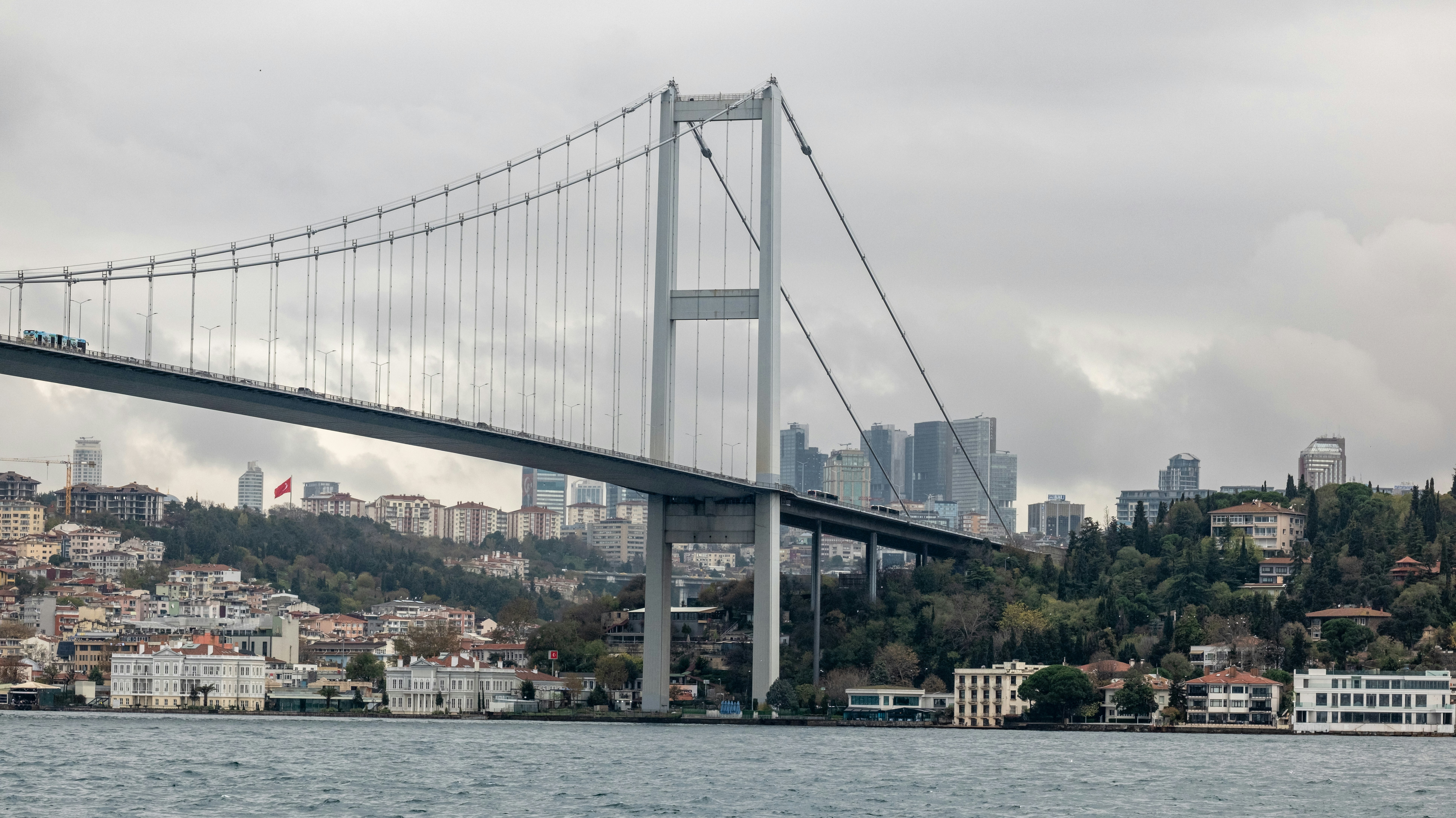 Suspension bridge over water with city buildings and green hills.