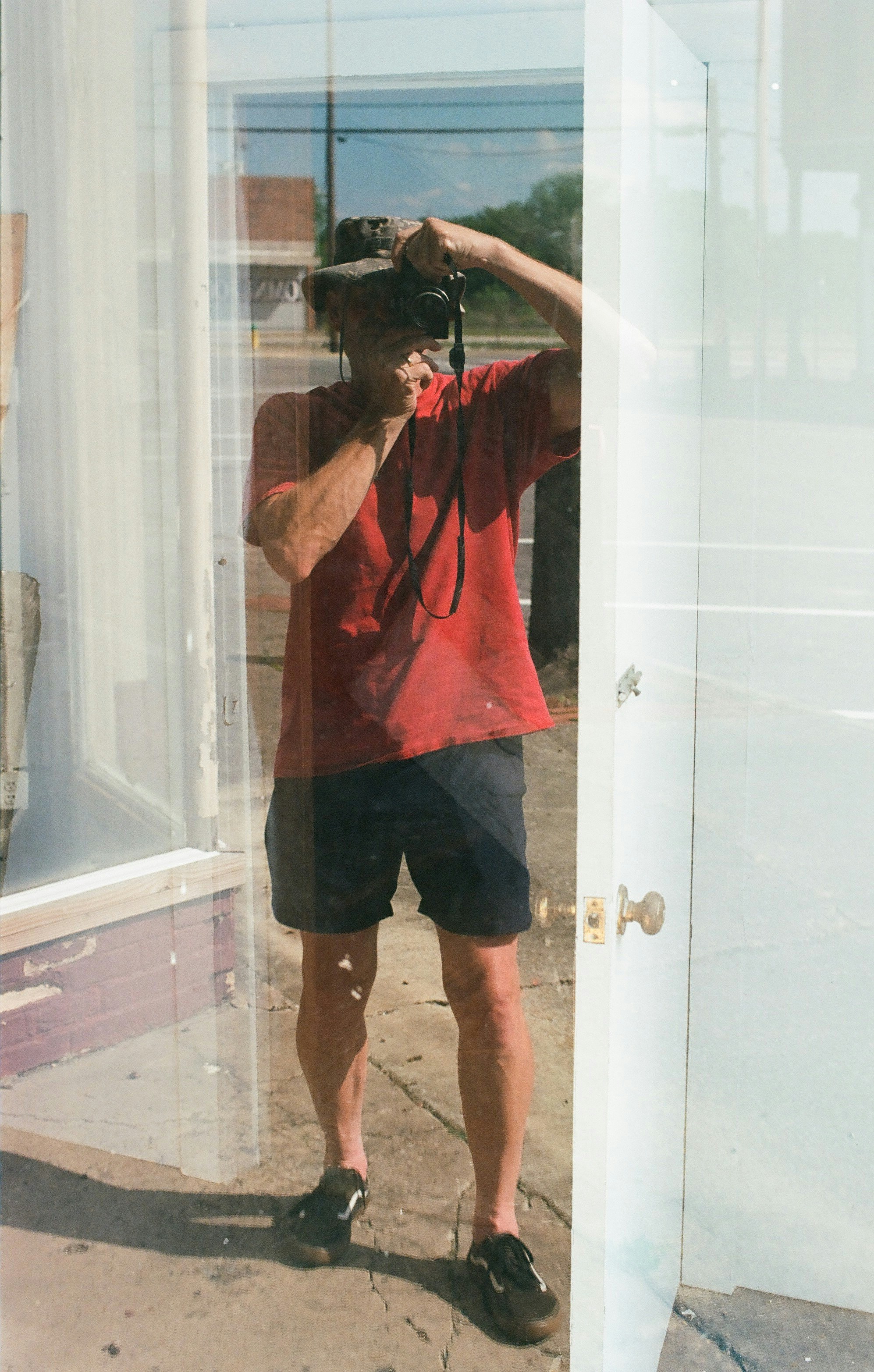 Self-portrait through a glass doorway: a sunlit photographer in a red shirt