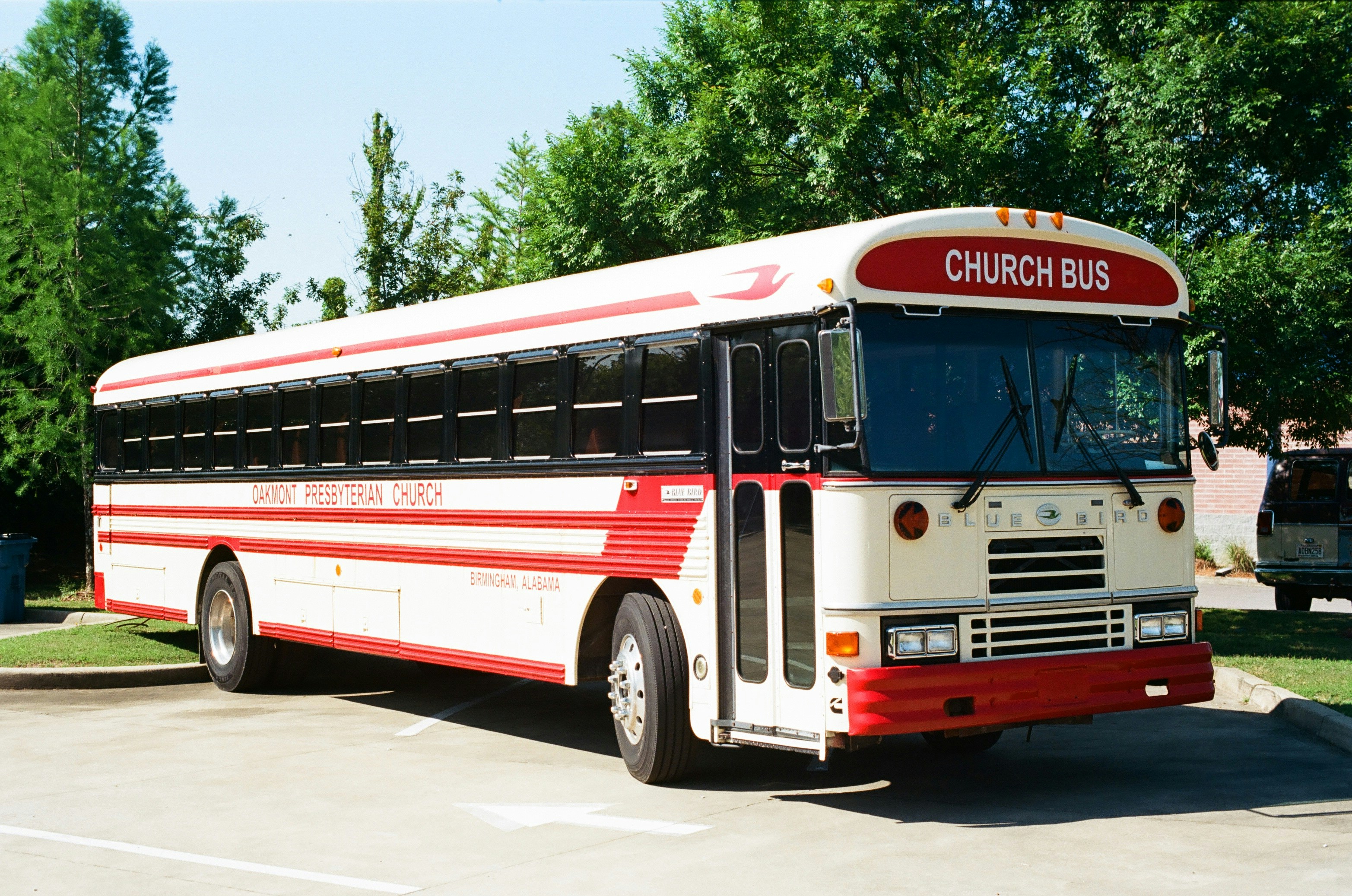 White Church Bus with Red Stripe Parked under Sunlit Trees