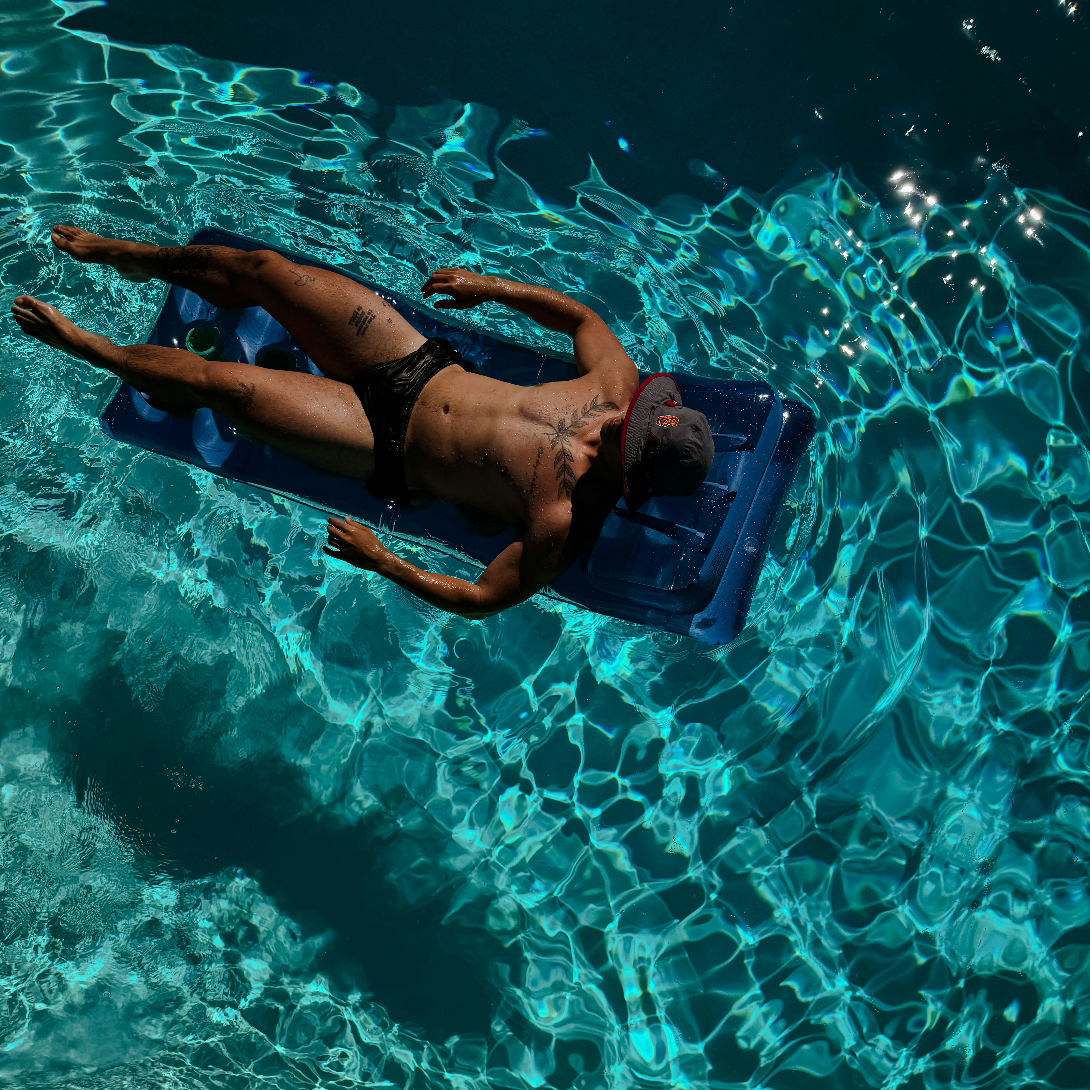 A swimmer floats face-down on a blue pool mat, sunlit turquoise water rippling around with clear reflections and shadowed edges.