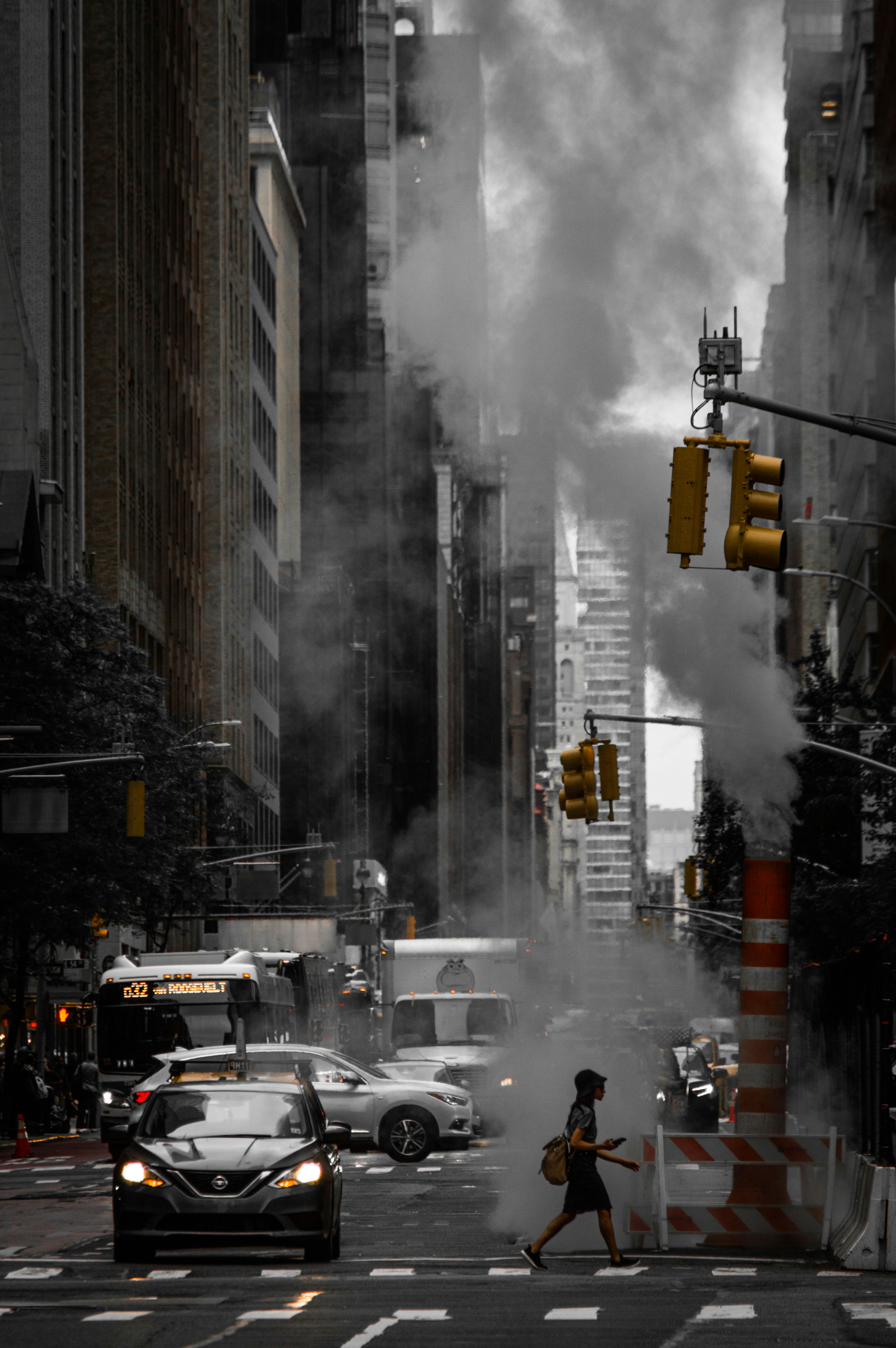 A gritty urban street scene with steam rising between buildings as a pedestrian crosses the road; traffic and construction barriers frame the foreground.