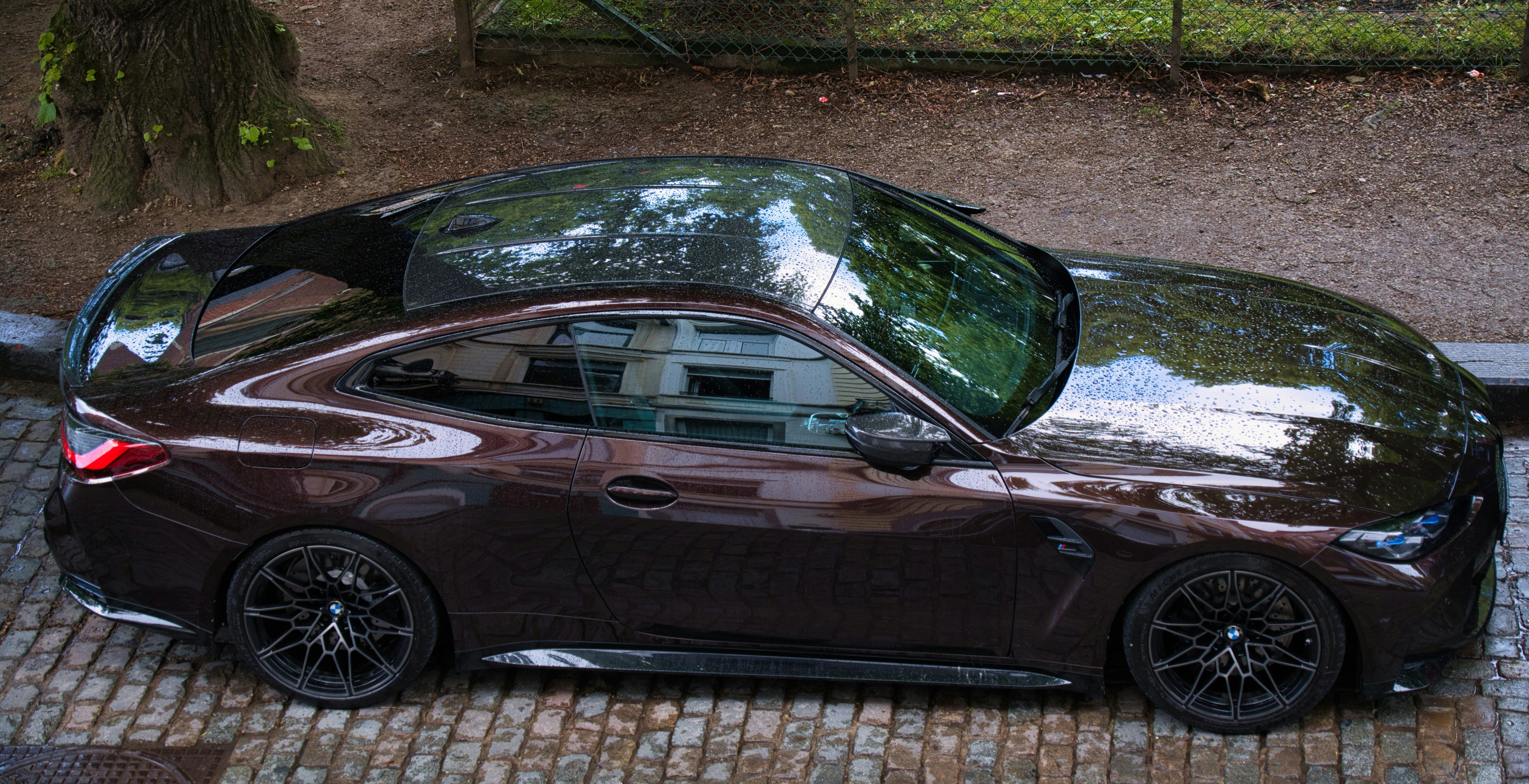 Dark metallic sedan captured from above on cobblestones, reflecting trees and sky; vehicle details and wheel design emphasized in a clean street scene.