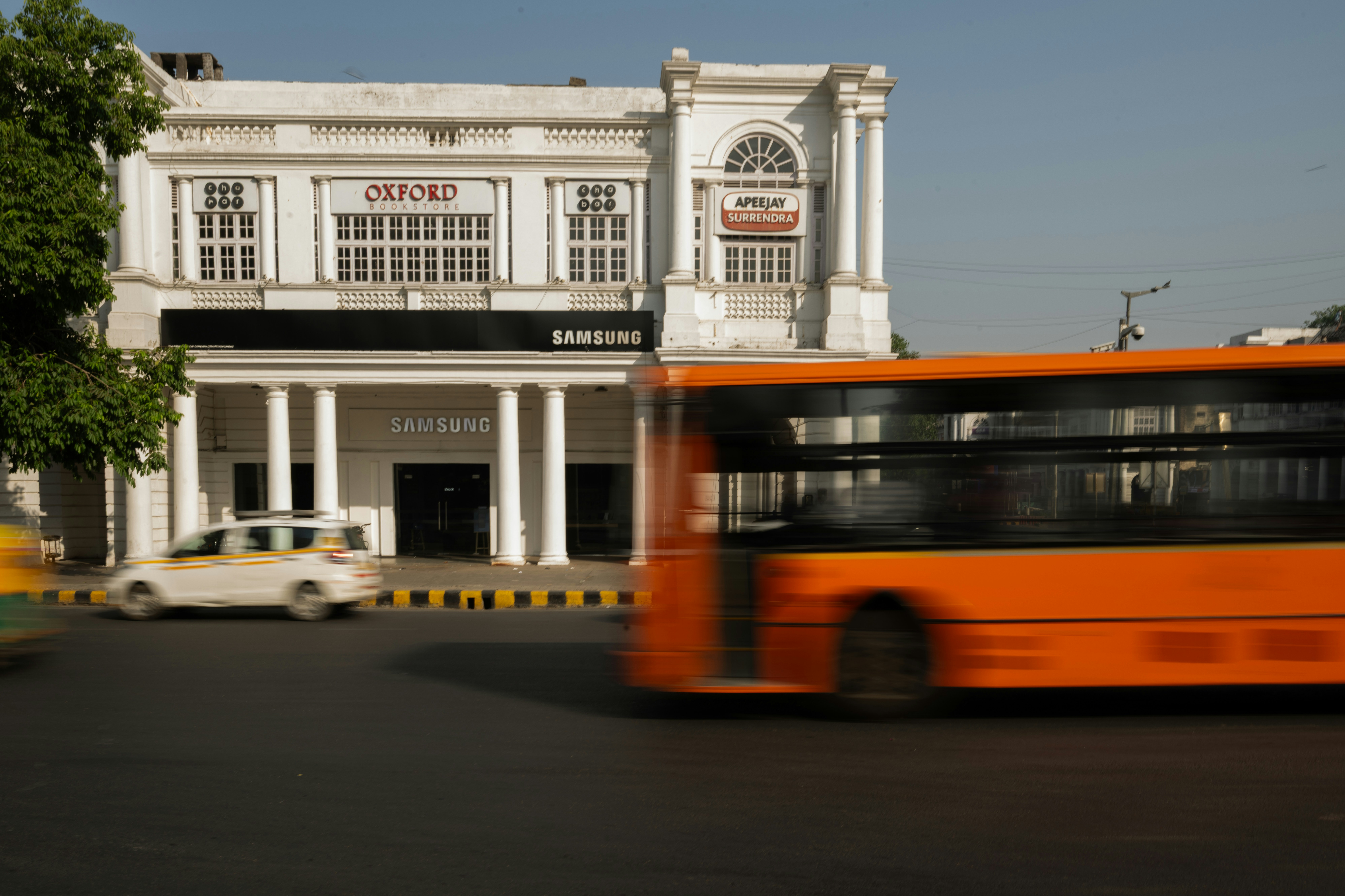 An orange bus blurs past a classic white building.