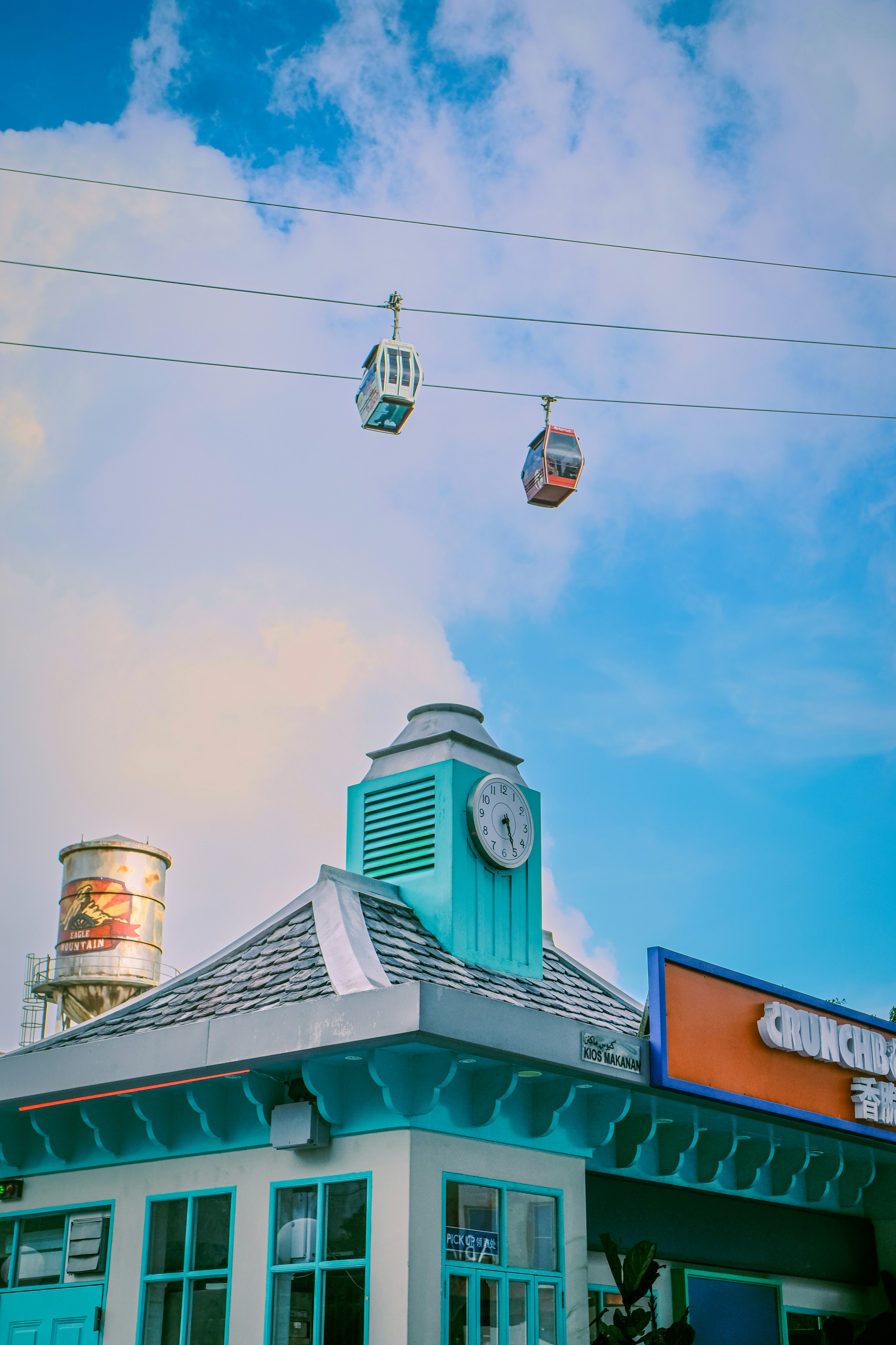 Skyway above teal storefront: two cable cars drift past a clock-topped building