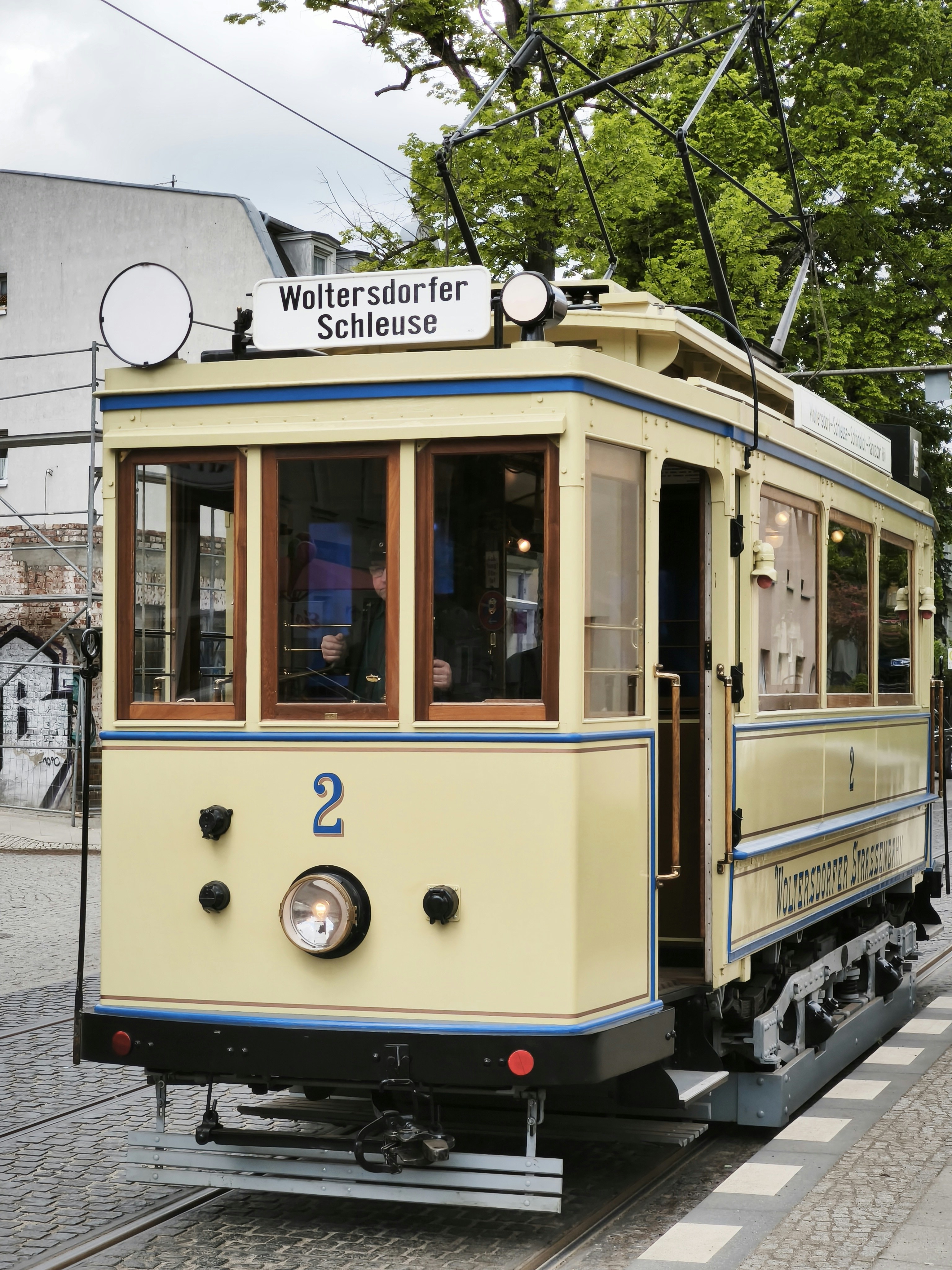 A vintage cream-yellow tram with blue trim sits on cobblestone tracks, its front face featuring a large headlamp and the number 2, while passengers are visible through the windows under a pale sky.