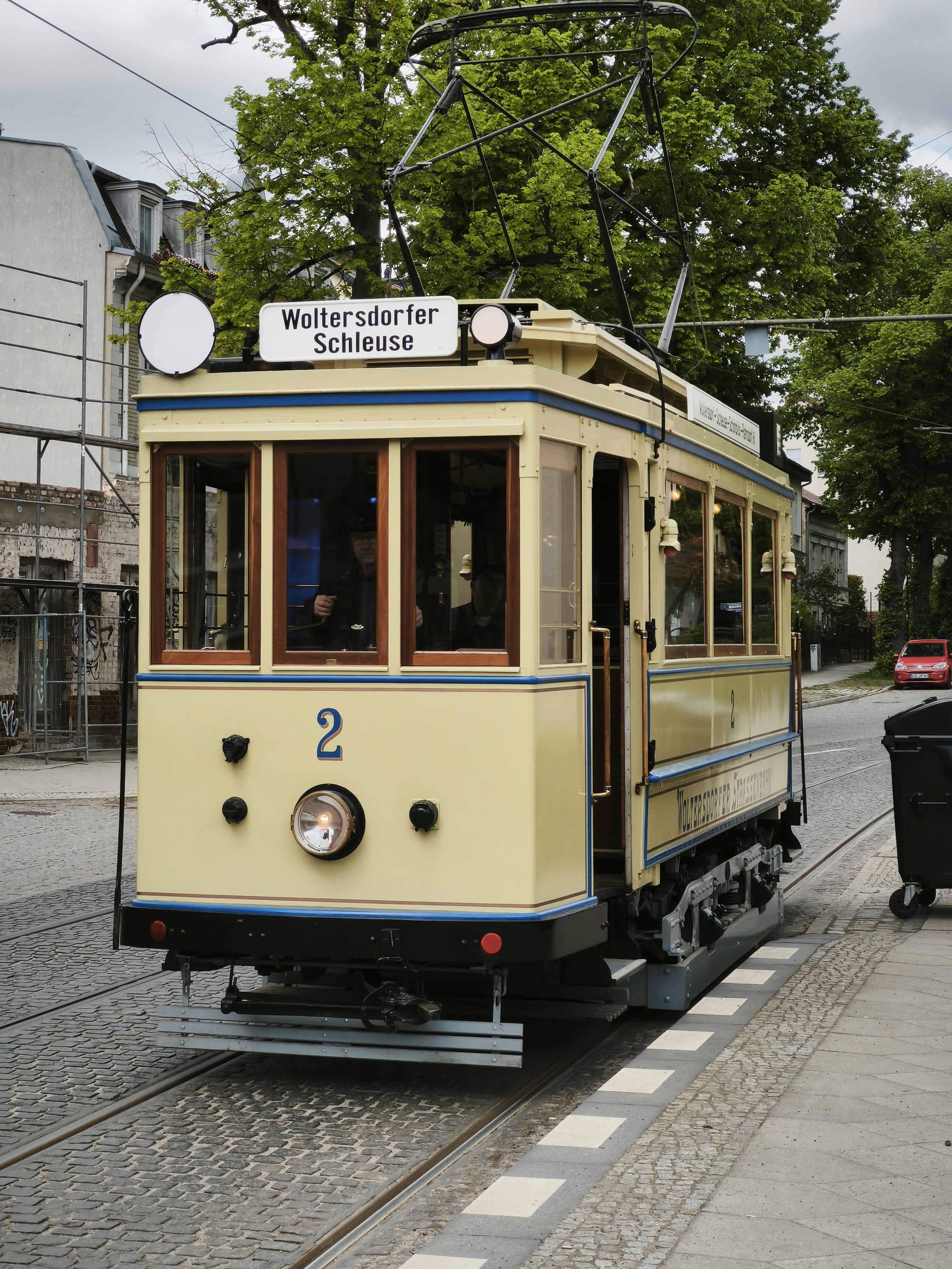 Cream-and-blue vintage tram gliding along cobblestone tracks with trees and buildings in the background; emphasis on the vehicle as the main subject, with a hint of its surroundings. The scene is captured as a documentary shot.