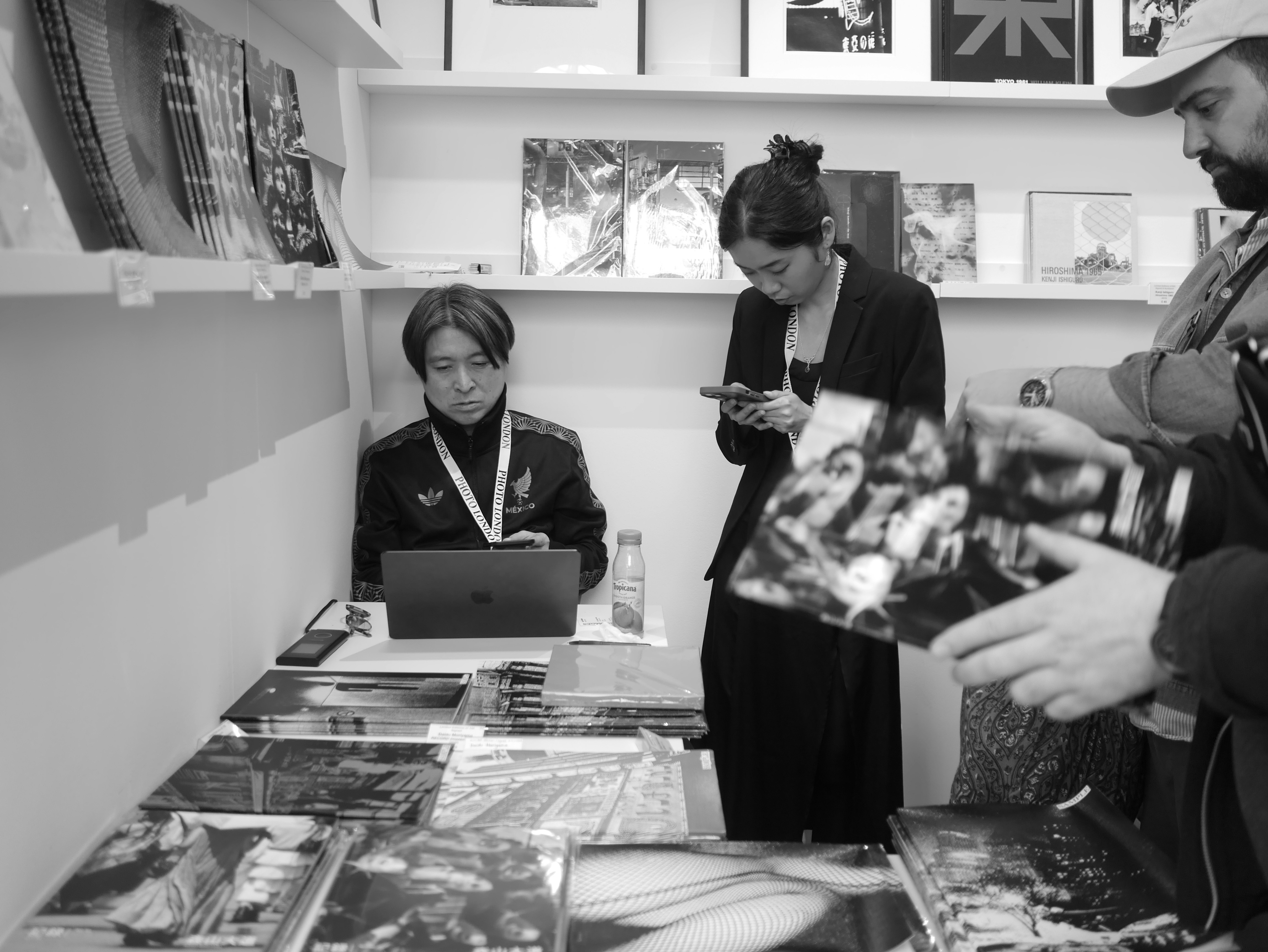 A man sits at a table with a laptop in a bookstore or gallery, while a woman and another man browse books and magazines around him. The scene emphasizes a small, lively space filled with printed materials and interaction.