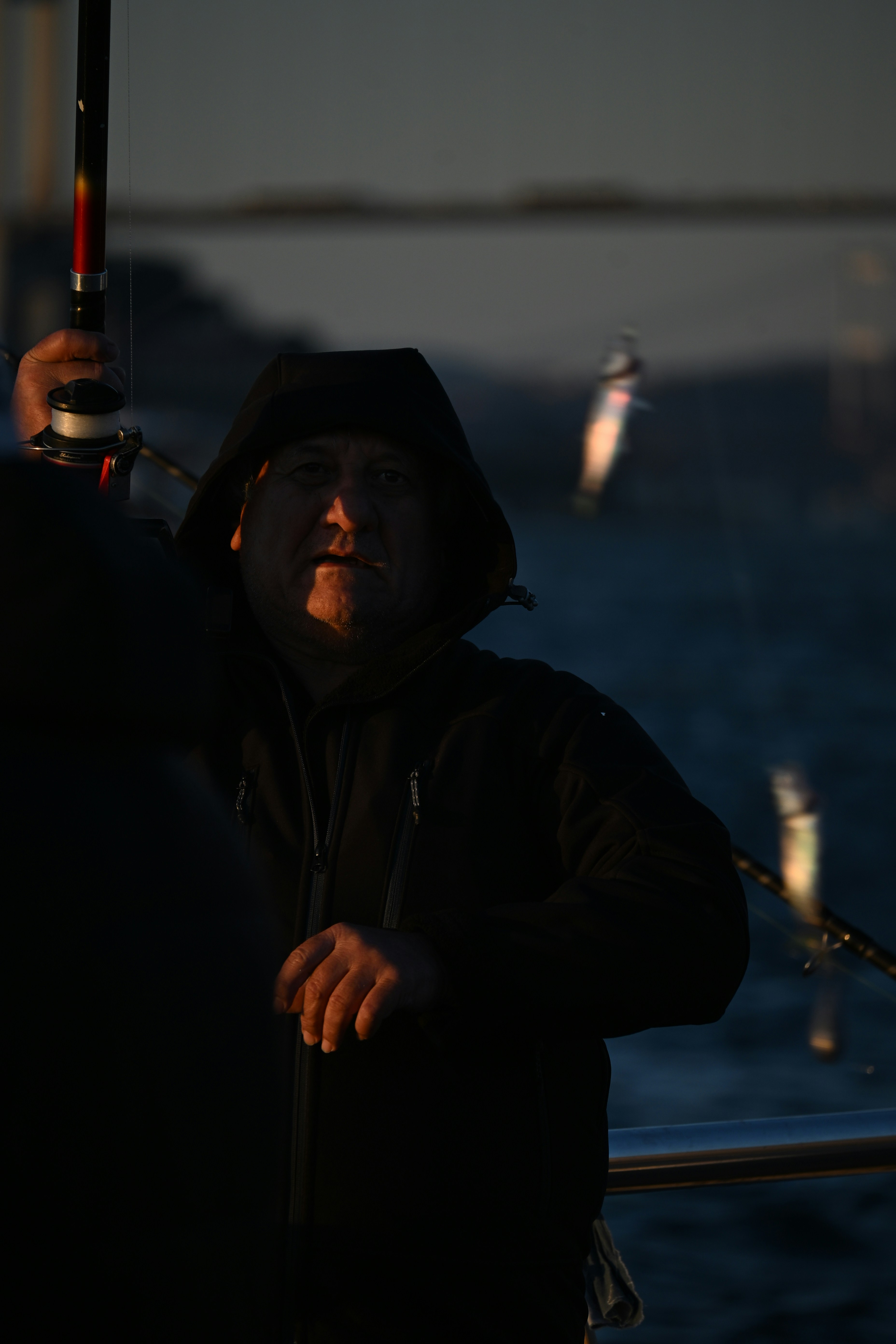 A solitary figure on a dimly lit boat deck, hood up against the chill, pierces the frame with a subtle presence against the sea. The scene captures a moment of quiet focus and maritime atmosphere.