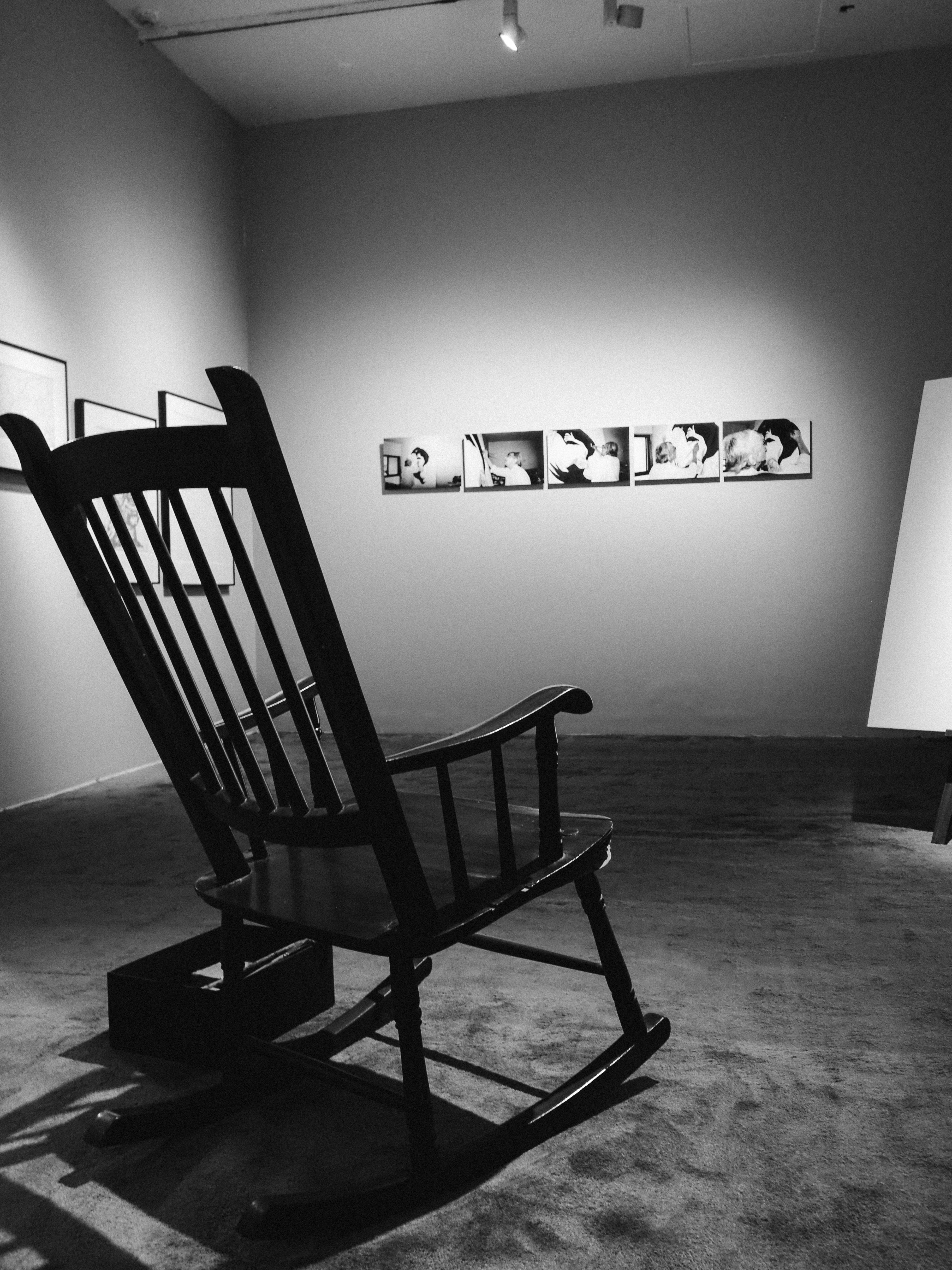 A solitary rocking chair sits angled in a dim gallery, with black-and-white tones and photo frames on the wall behind it.