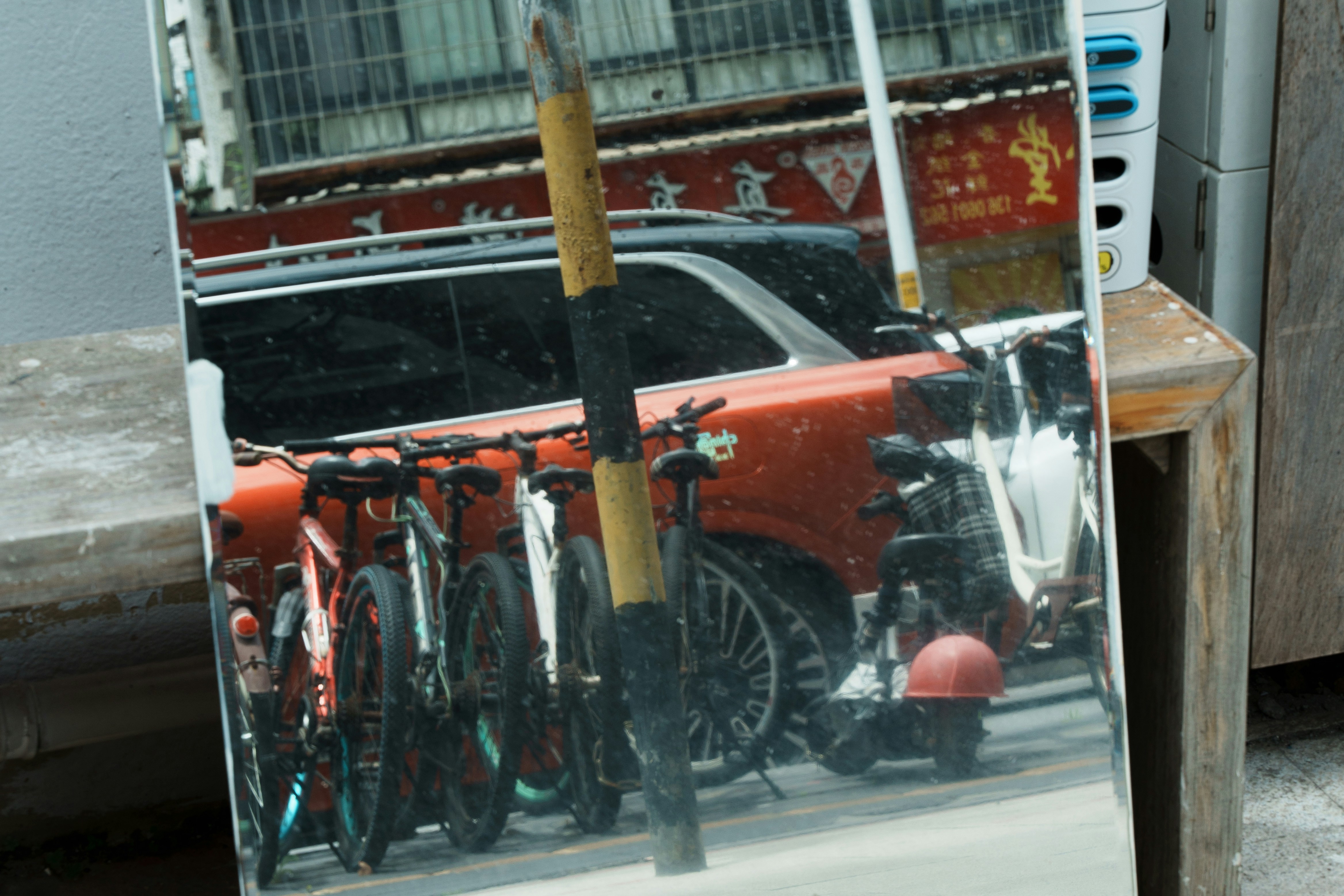 Reflected Street: a mirrored scene of bikes and a red car on a rainy storefront
