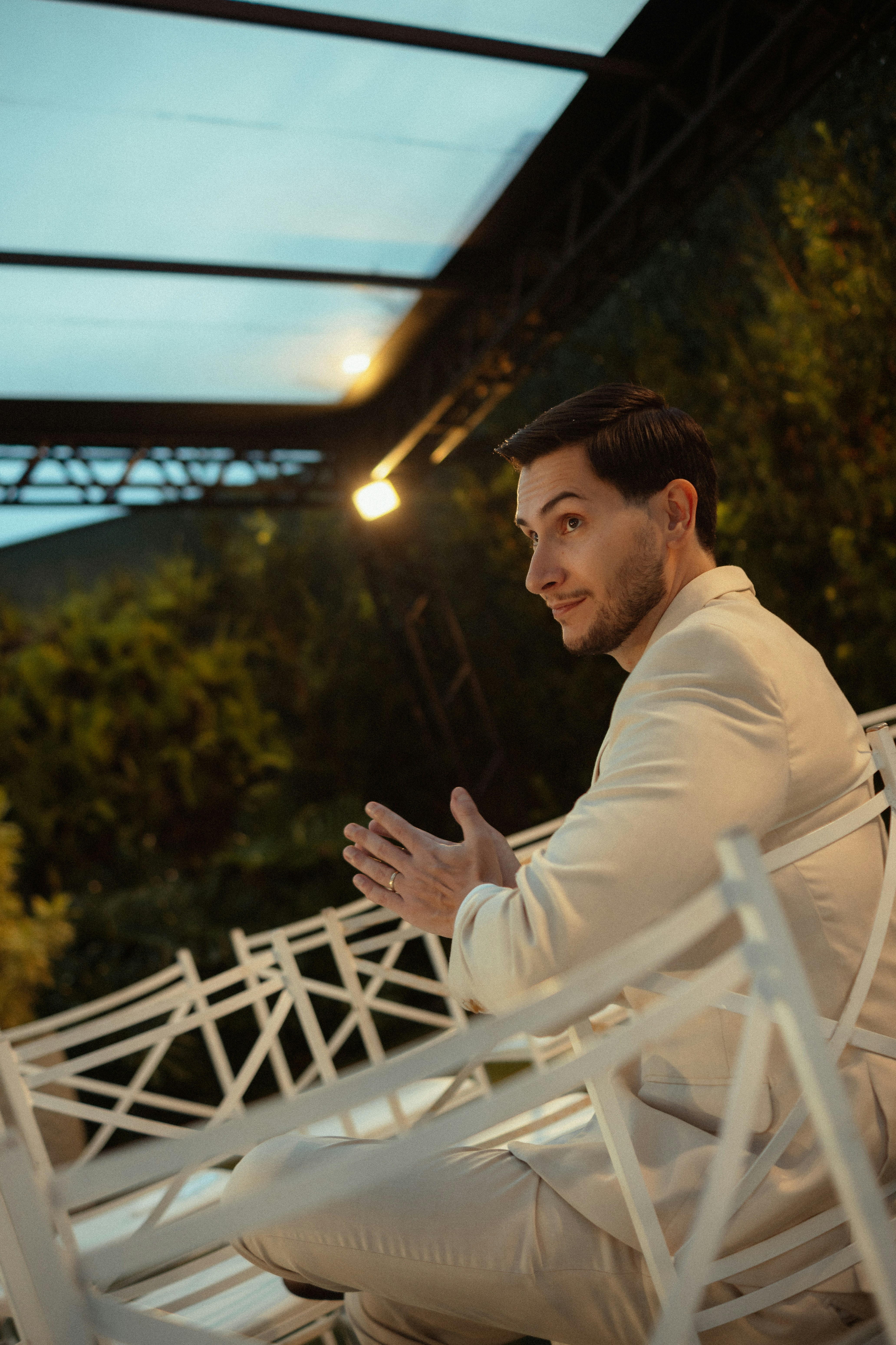Man in light suit sitting outdoors, hands clasped, smiling.