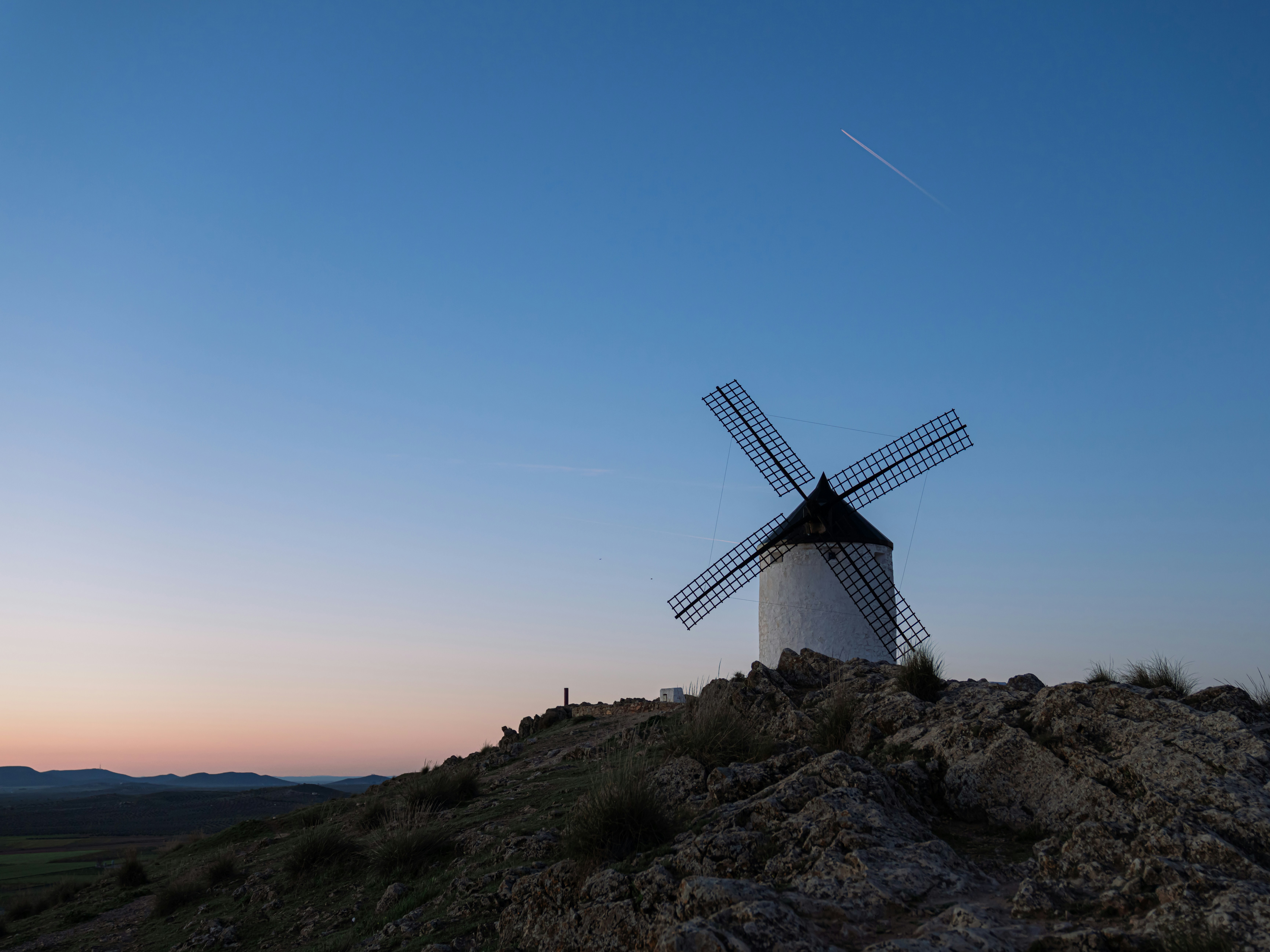 Low-angle view of a windmill perched on a rocky hill at sunset, with the horizon glowing orange and the sky fading to blue.