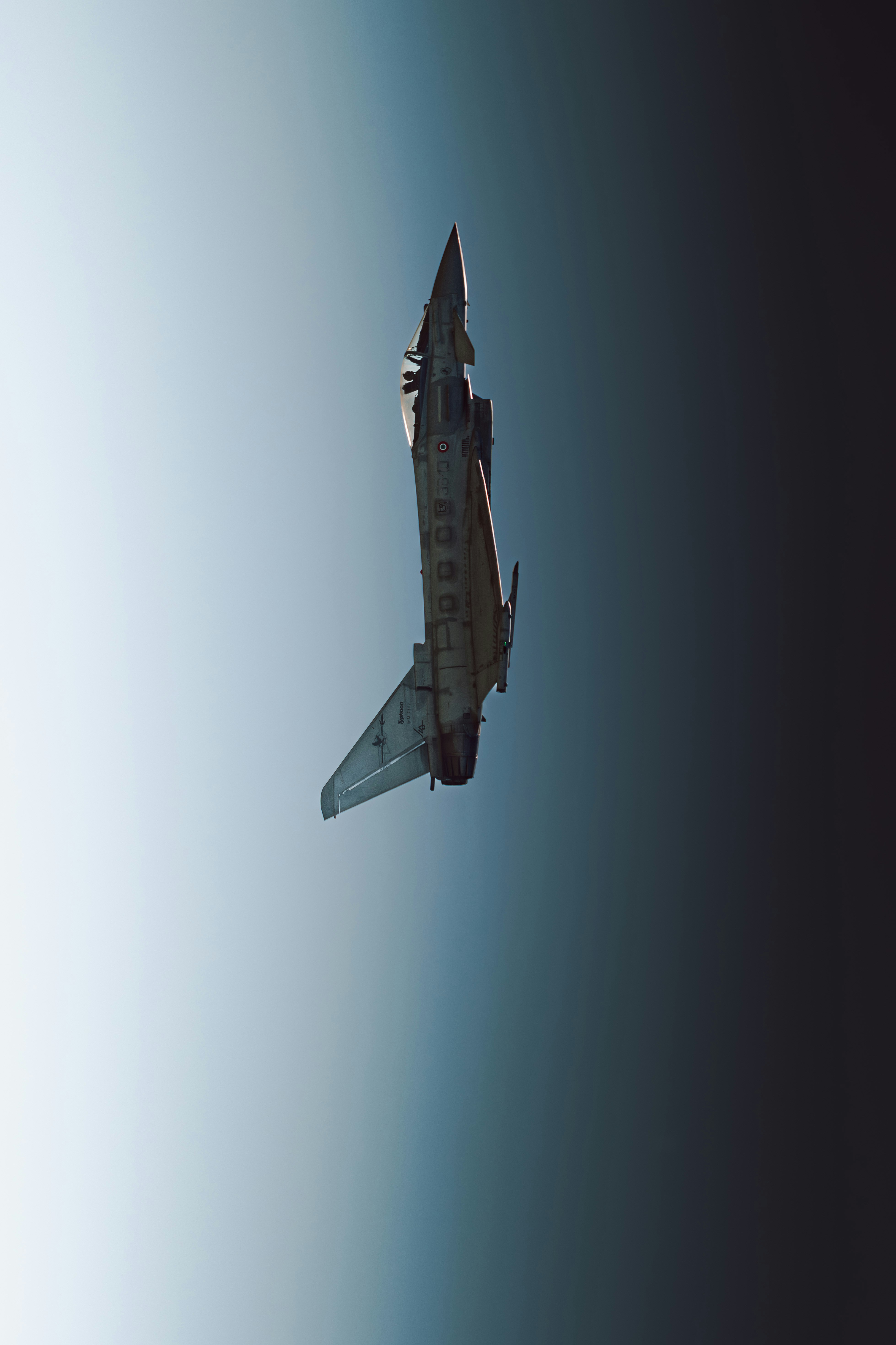 Military jet inscrutably ascending through a deep gradient sky, captured from a dramatic underside perspective. The scene emphasizes the aircraft’s silhouette and engineering detail without background clutter.