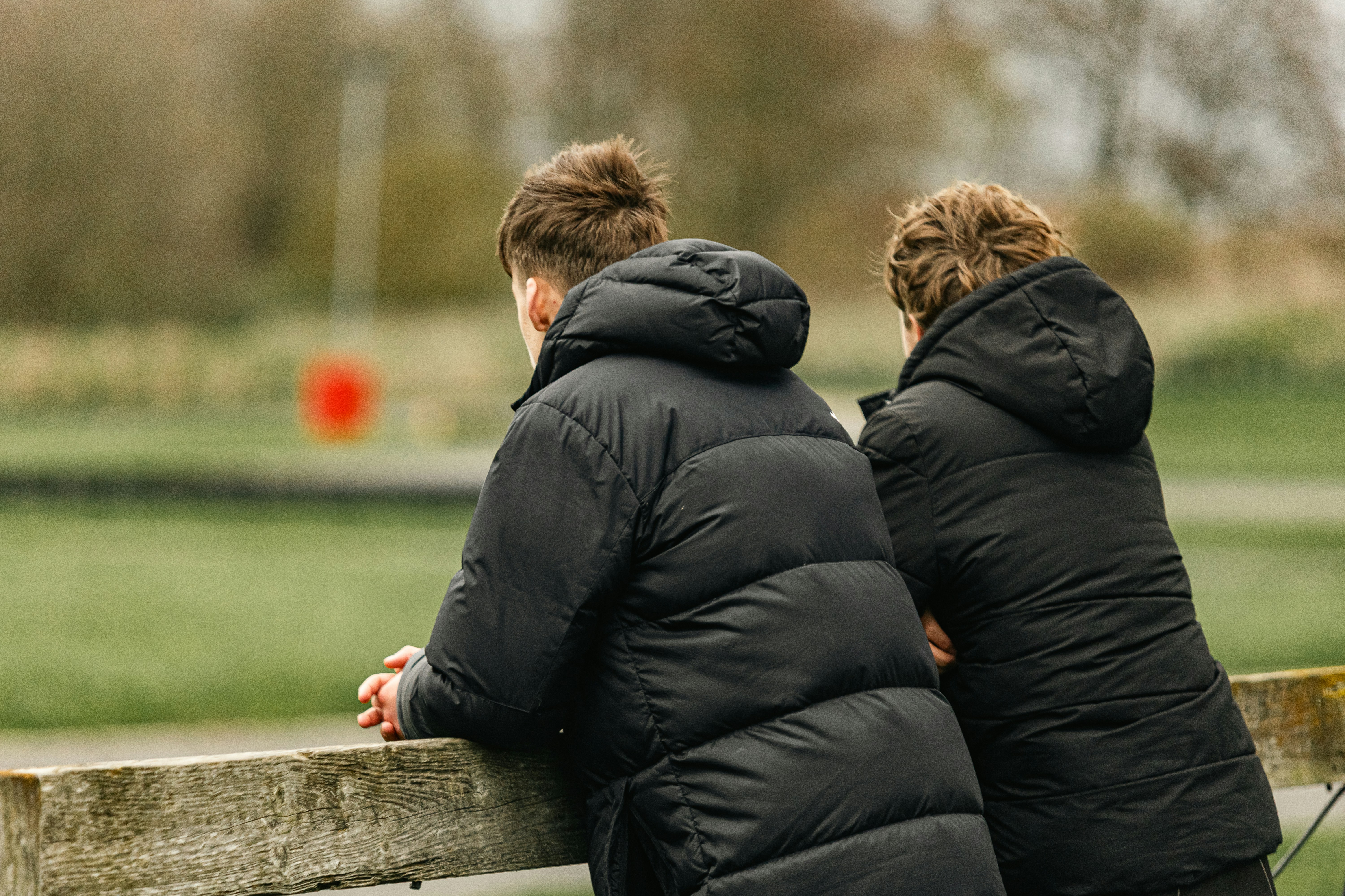Two Friends in Matching Parkas Lean on a Weathered Fence
