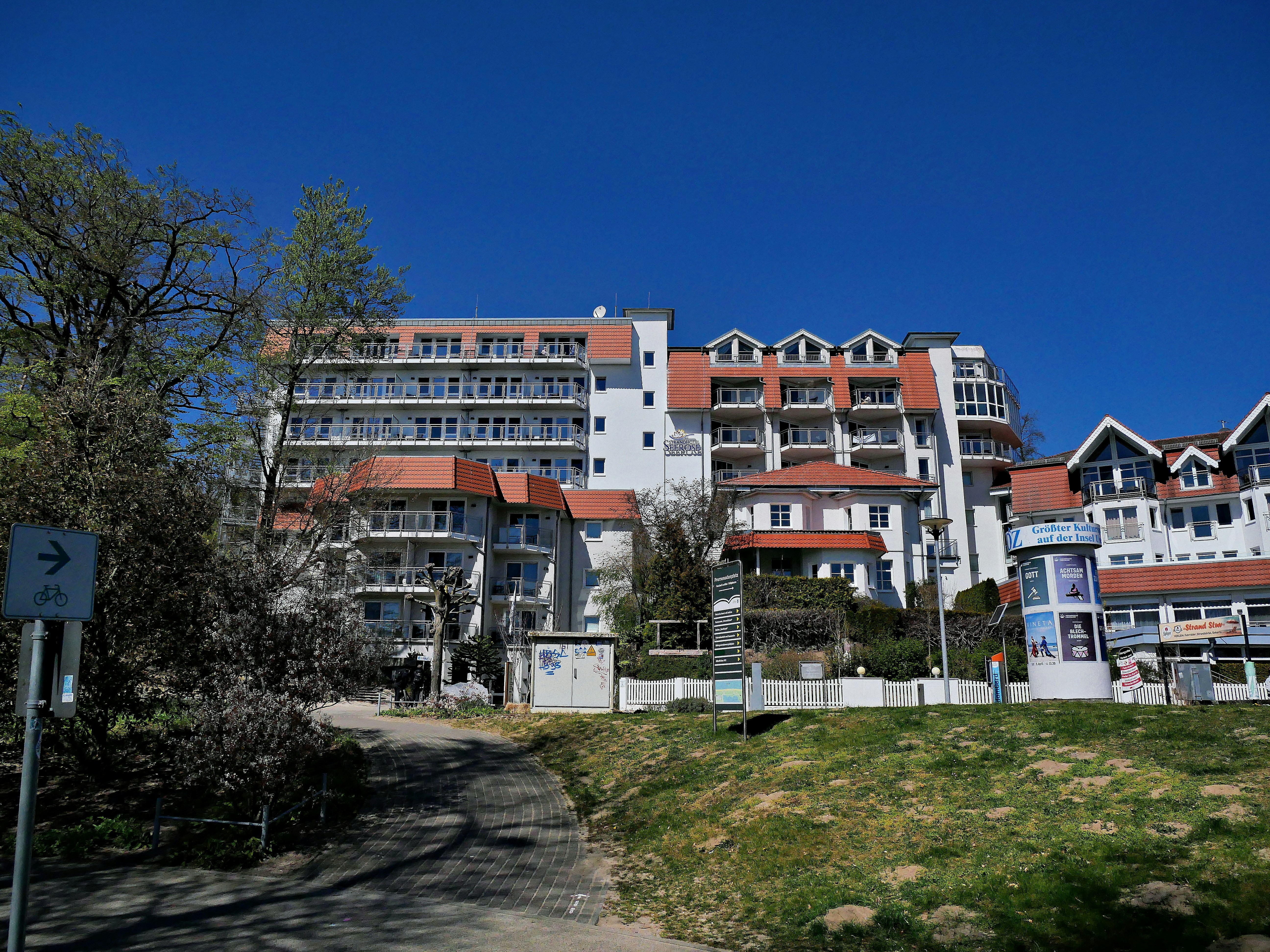 Wide shot of a multi-storey residential complex under a deep blue sky, with a sunlit lawn foreground and a curved pathway. The scene emphasizes the architecture and surrounding greenery.