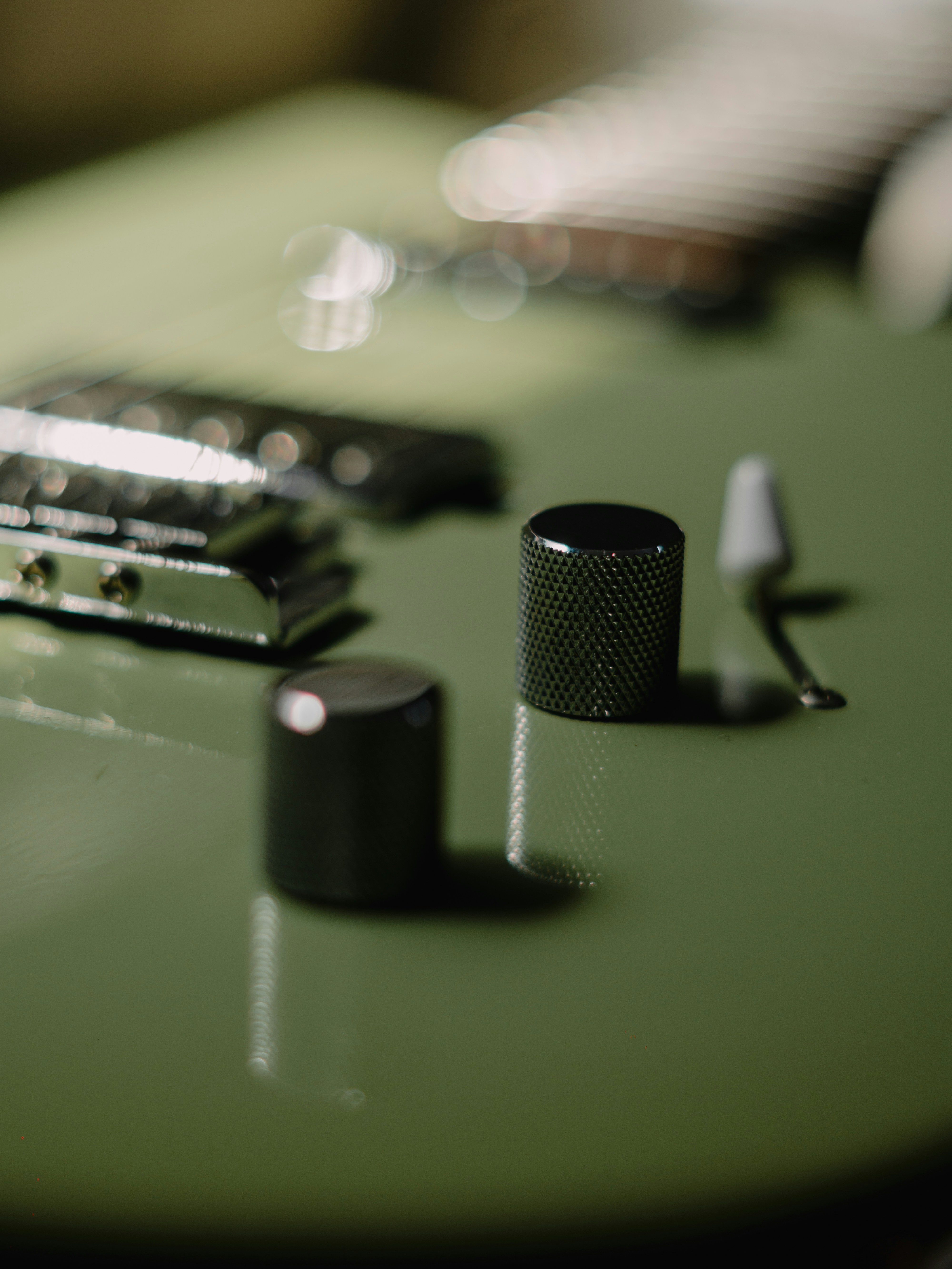 Close-up view of a green electric guitar focusing on the control knobs and bridge hardware, with soft bokeh and shallow depth of field to emphasize texture and metallic details.
