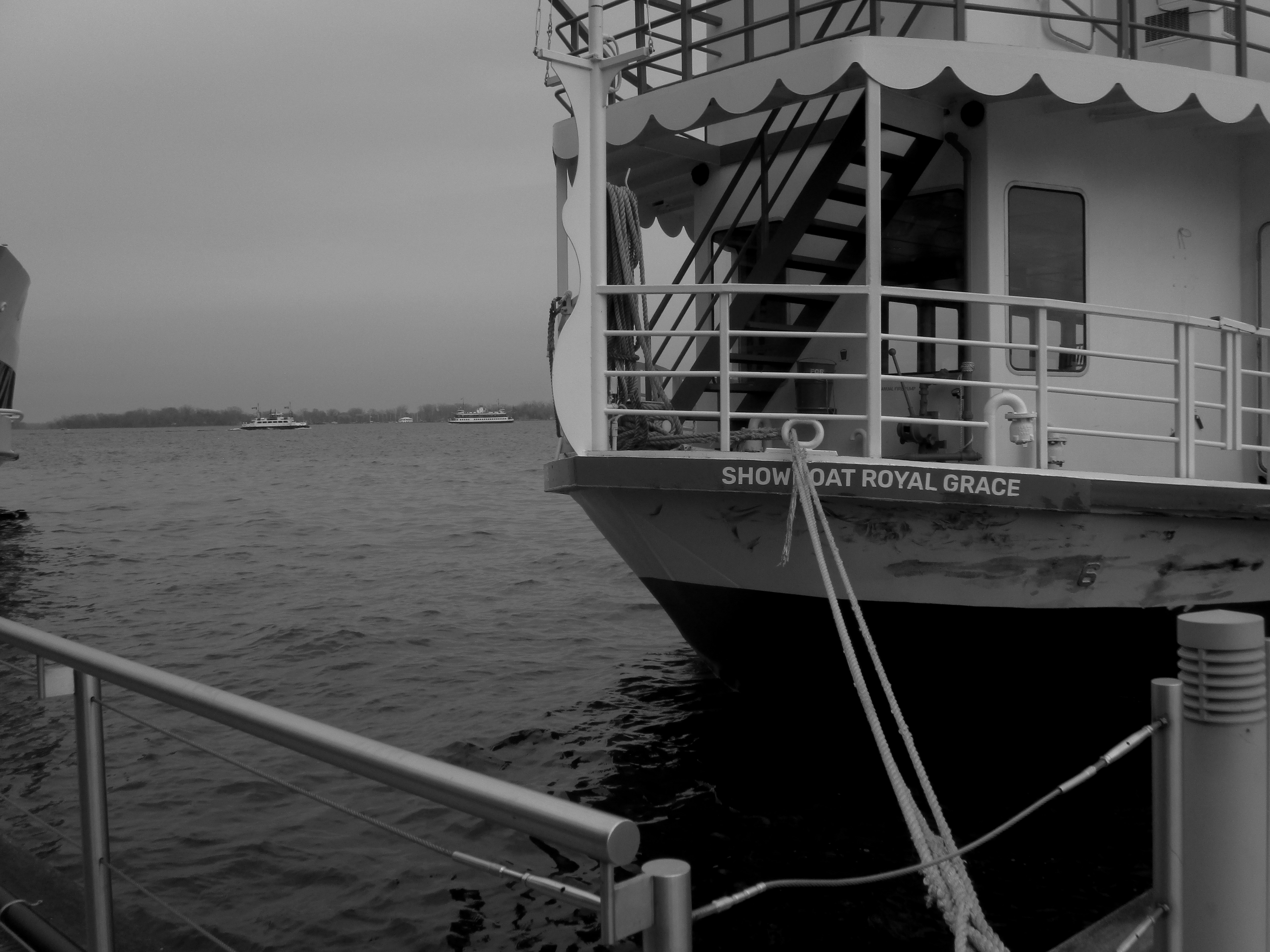 Muted grayscale scene of a docked boat with visible railings and ropes along the water, highlighting the vessel’s stern and deck details.